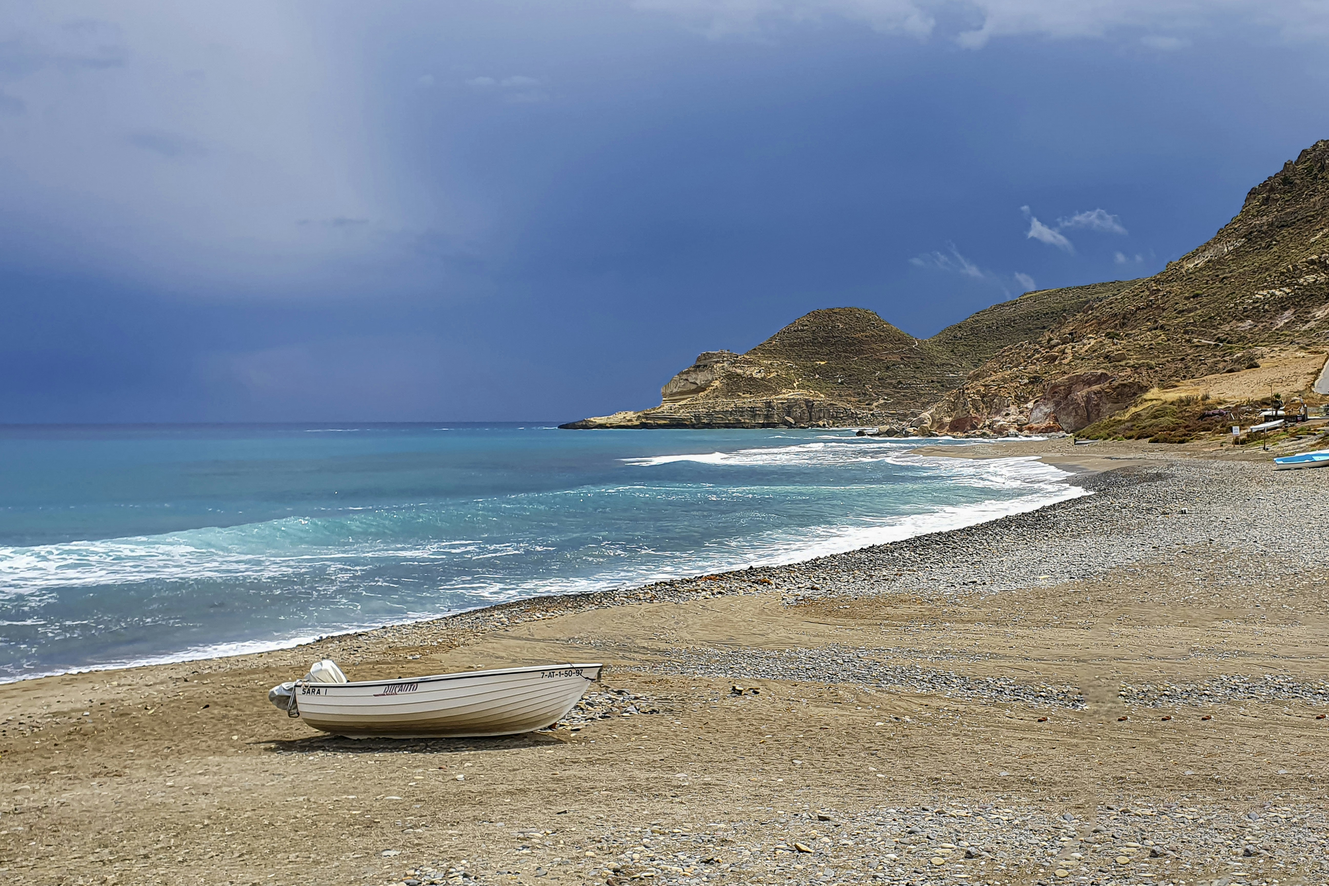 Small boat rests on a pebbled beach with turquoise waves under a moody sky.
