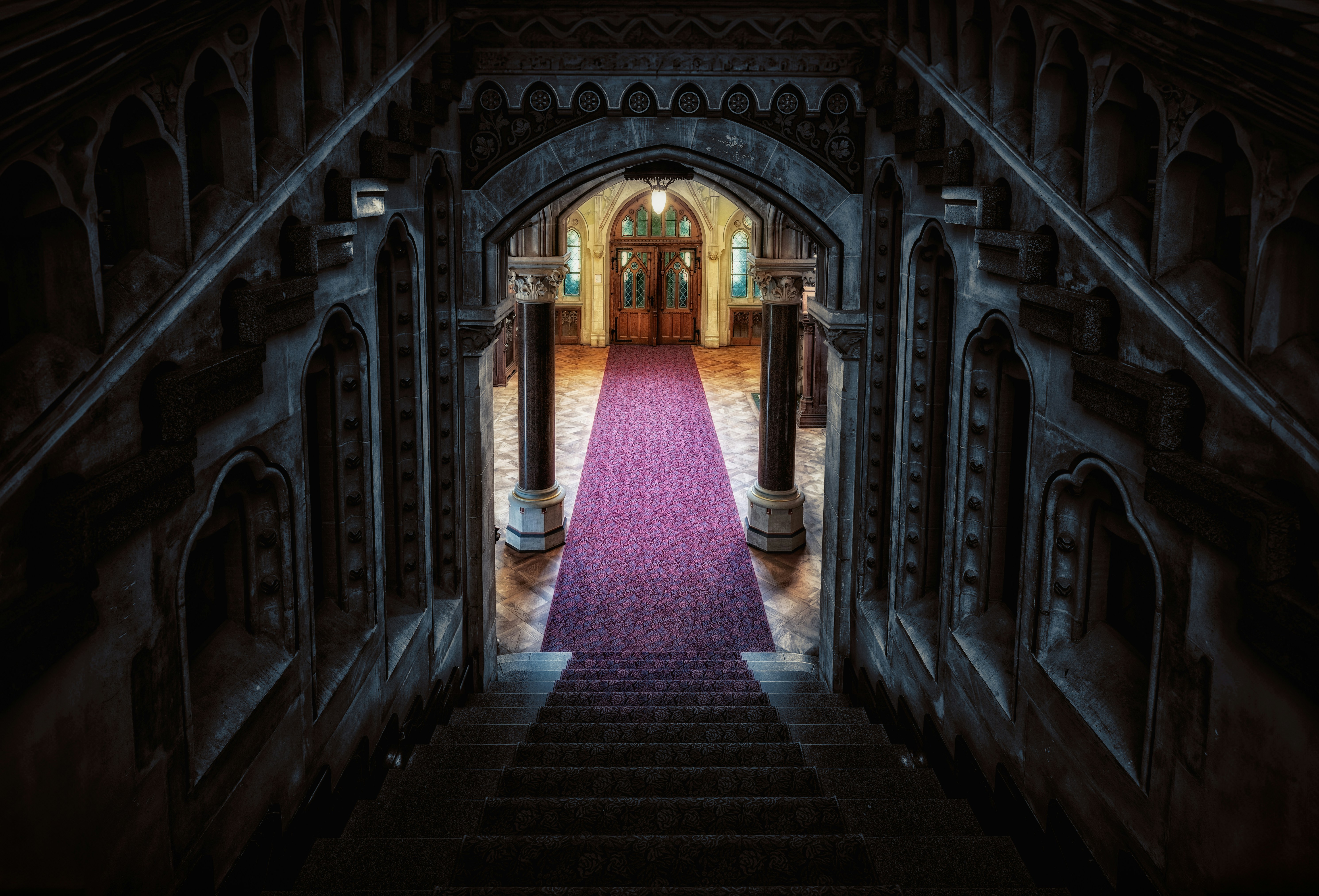 Dimly lit stone staircase framed by arches and columns leads toward a warmly lit doorway at the far end. A rich violet carpet runs down the central path, drawing the eye to the glow beyond.