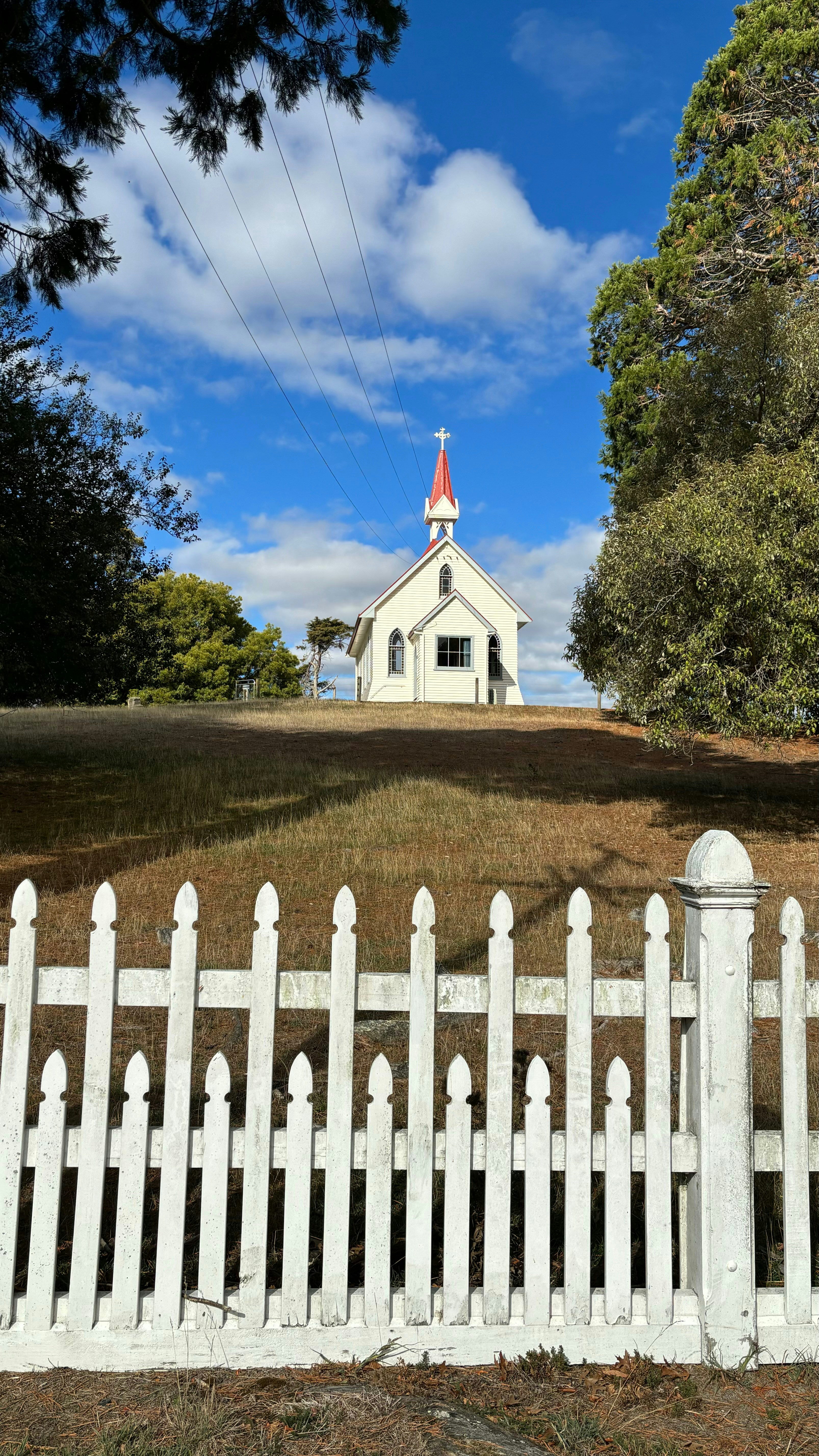 A small white church with a red steeple sits on a sunlit grassy hill, framed by a white picket fence. The blue sky and surrounding trees emphasize a tranquil rural scene.