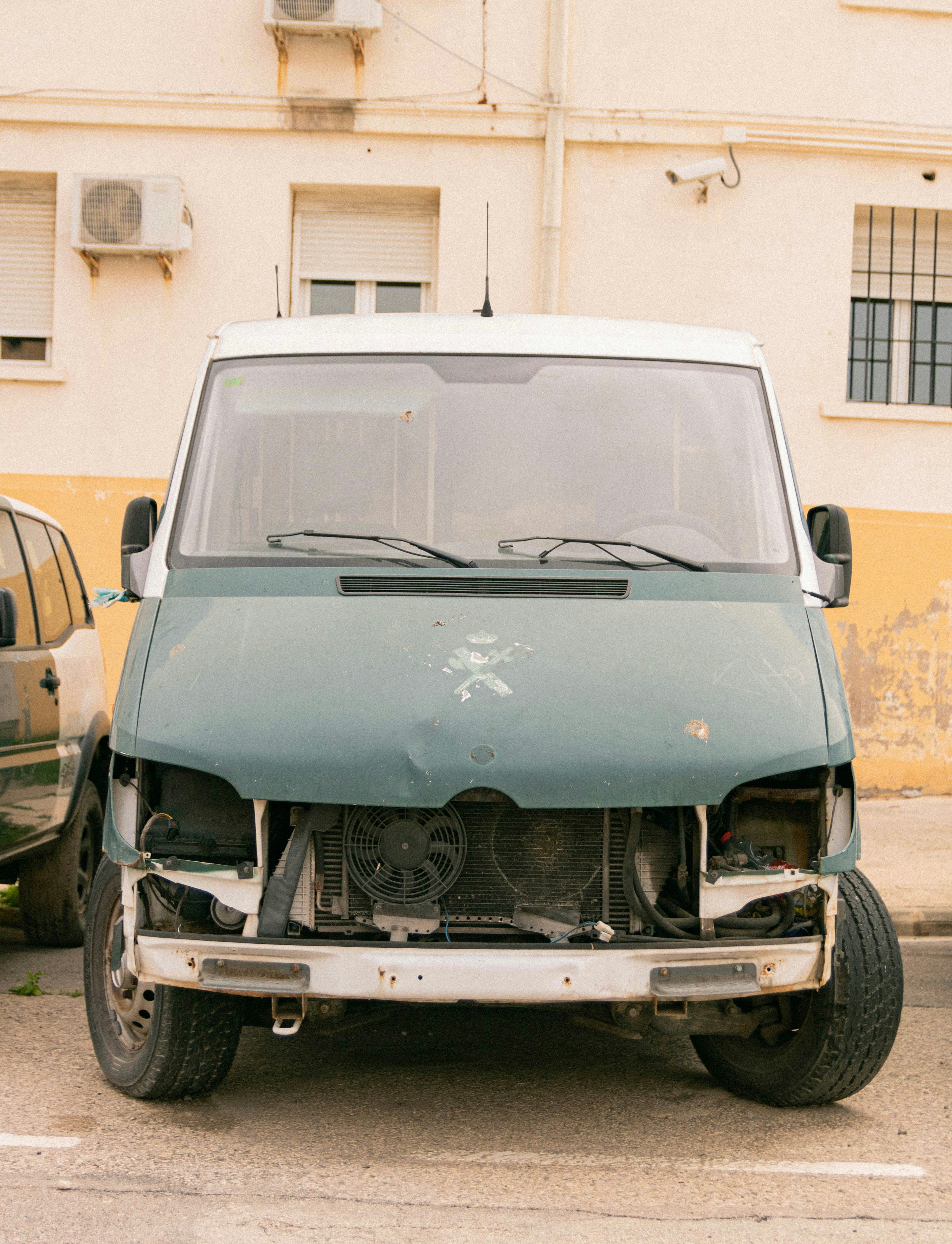 Front-Row Reveal: a weathered green van with exposed radiator in a sunlit urban lot