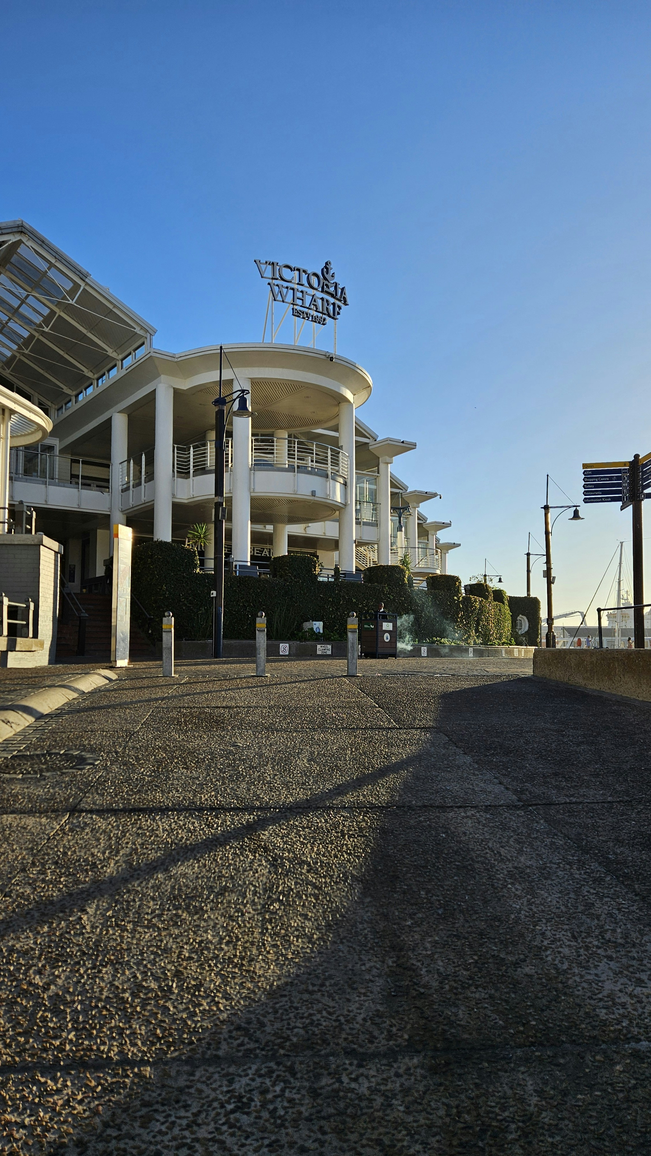 Sunlit promenade beside a curved, white, multi-level building labeled Victoria Wharf, with a marina and clear blue sky in the background.