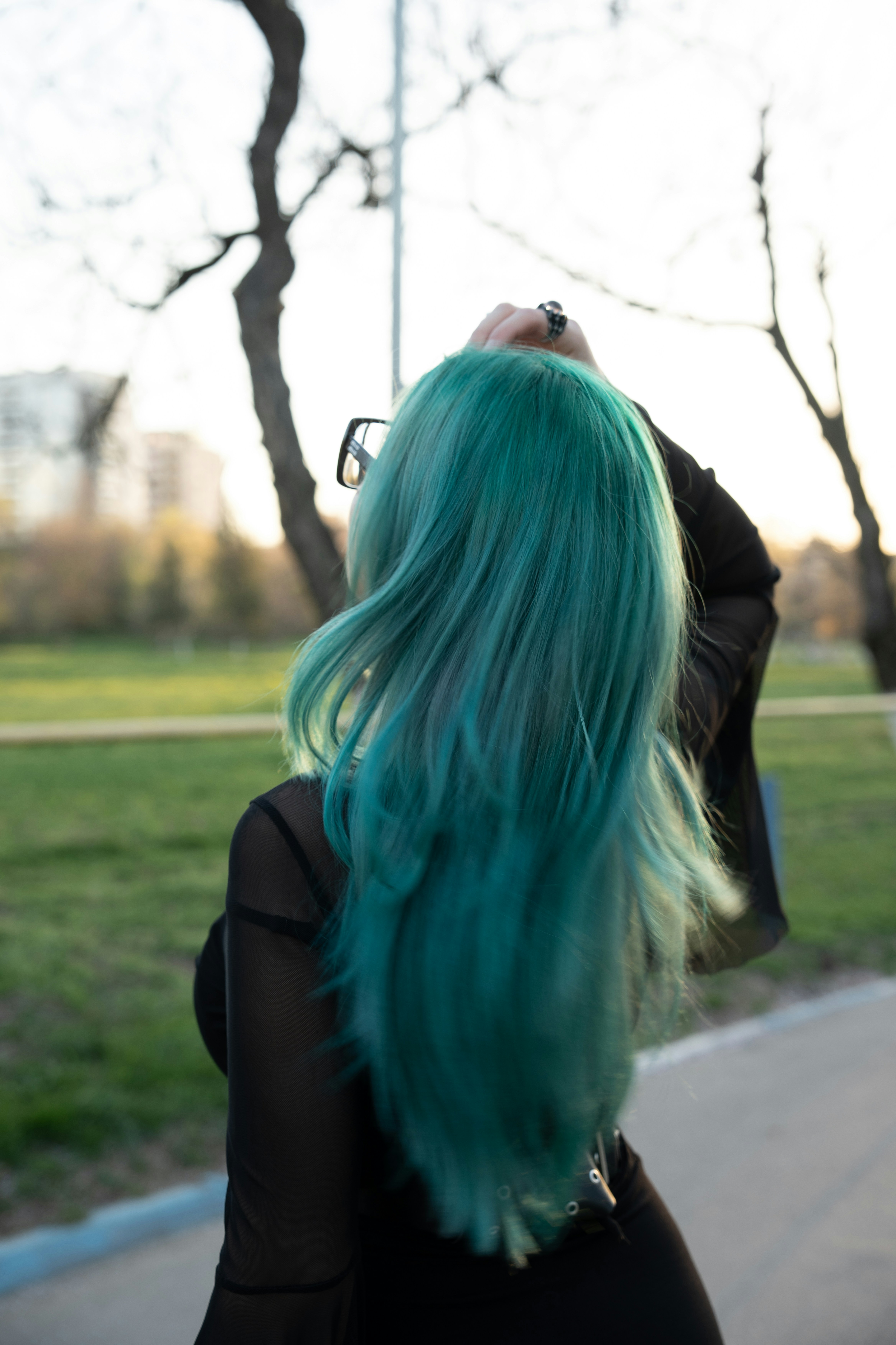 Back view of a person with teal hair, raising an arm in a sunlit park. The shallow depth of field keeps the background soft while the vibrant hair draws focus.