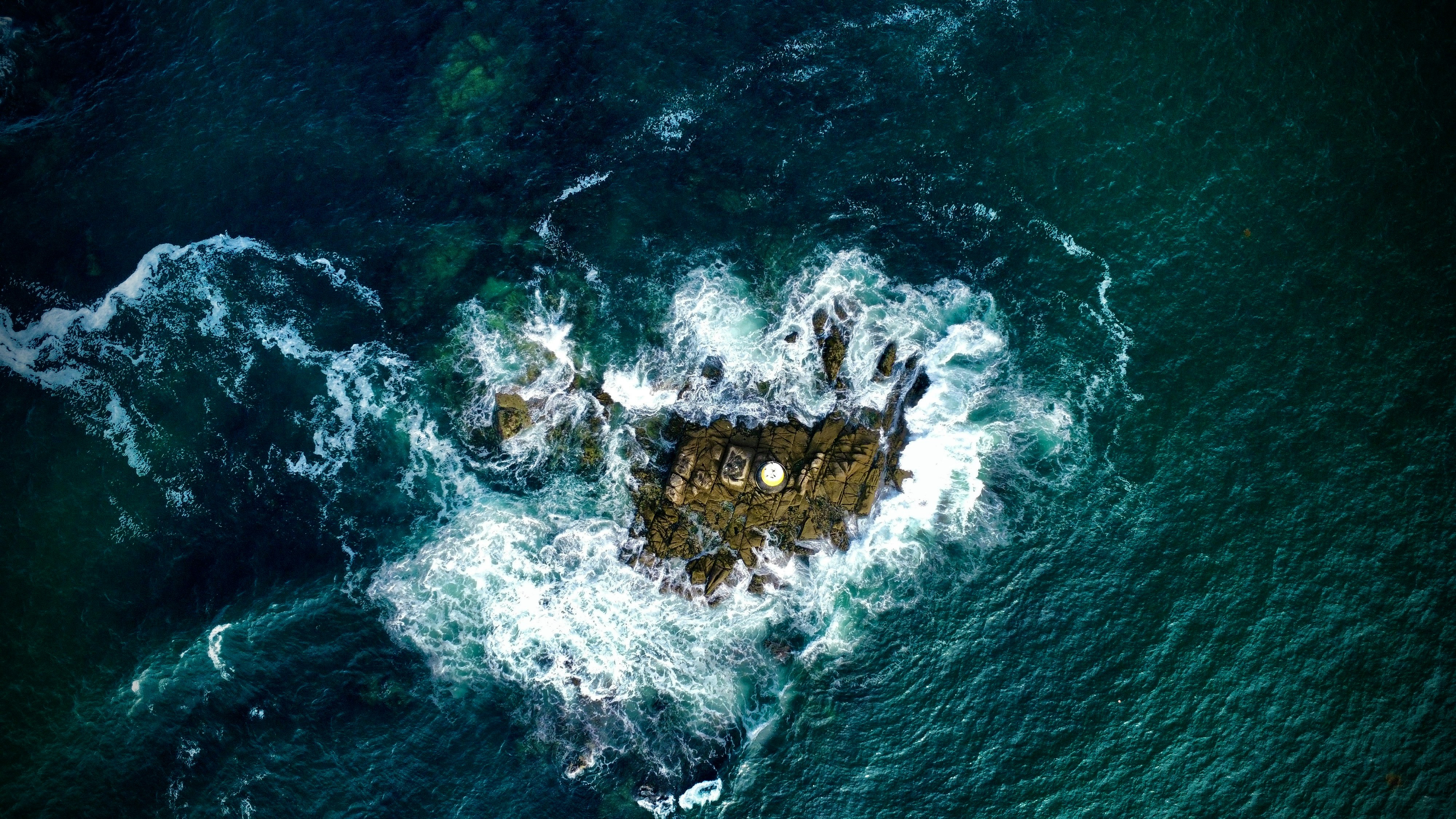 Waves crashing over a solitary rock formation surrounded by deep blue ocean waters.