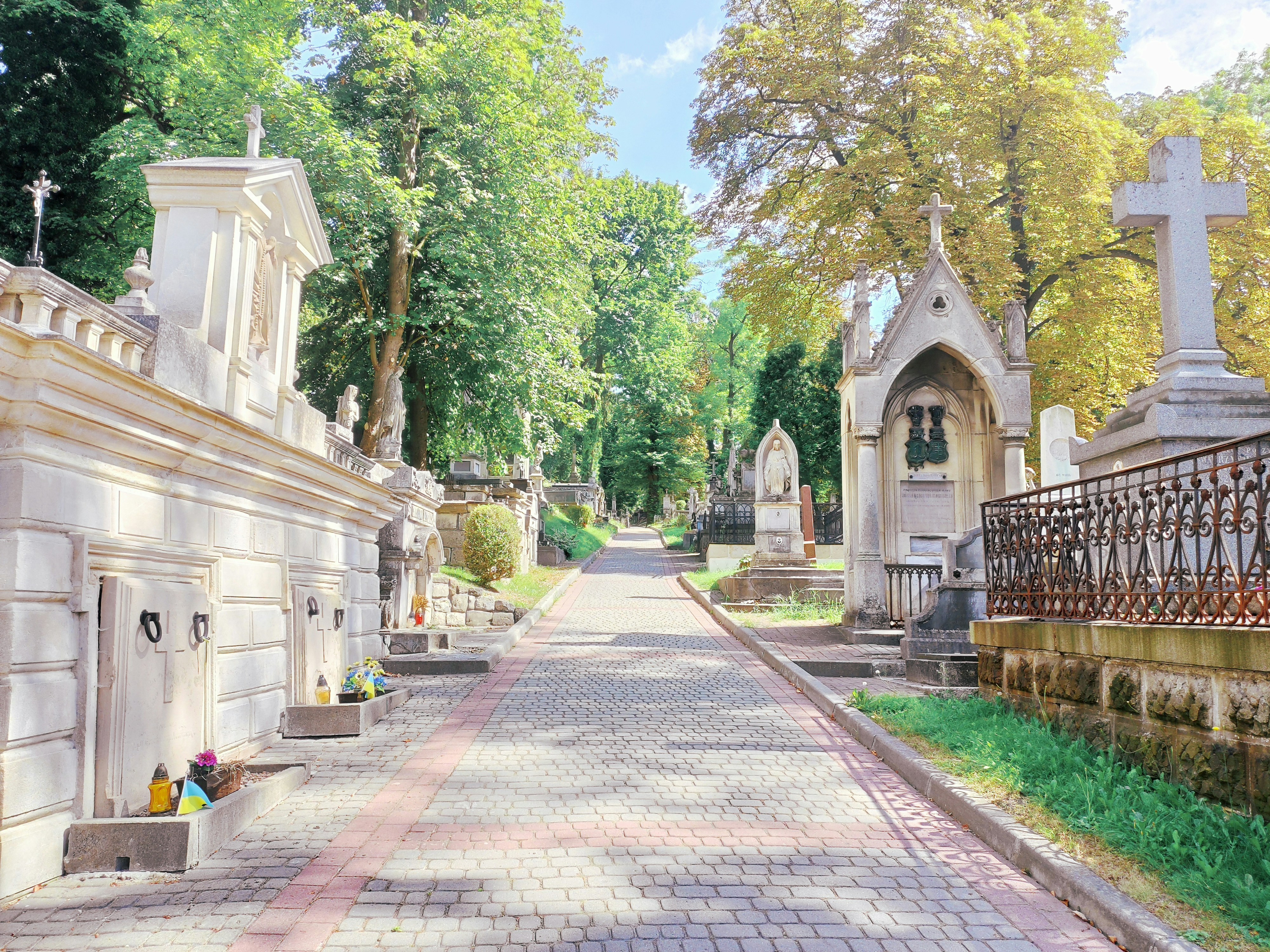 Quiet pathway lined with ornate tombs and lush trees, leading into a tranquil cemetery. The scene captures a sense of history and reflection.
