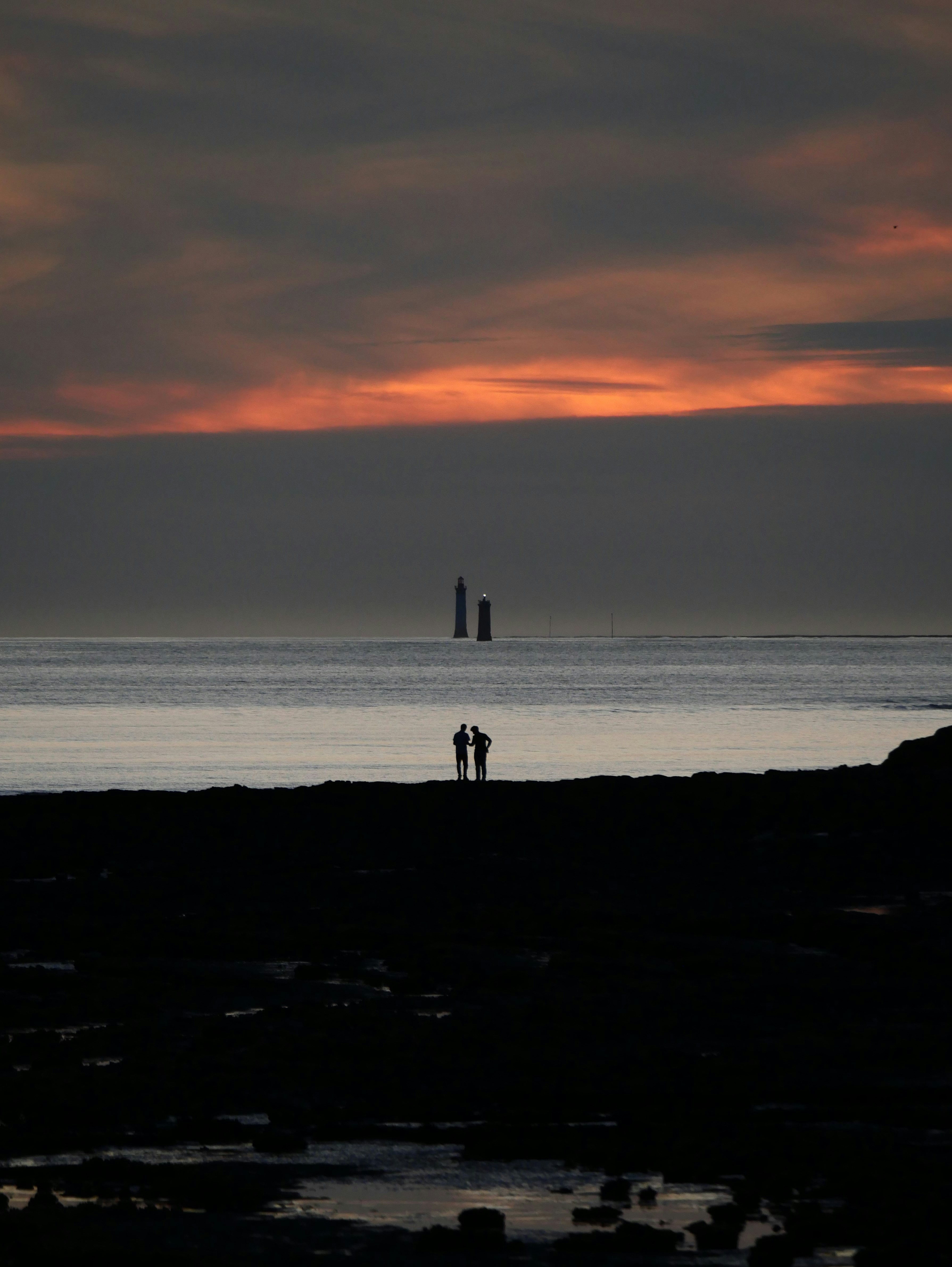 Seascape at sunset with two distant lighthouses on the horizon and silhouettes near the shore. Calm water reflects the warm sky.