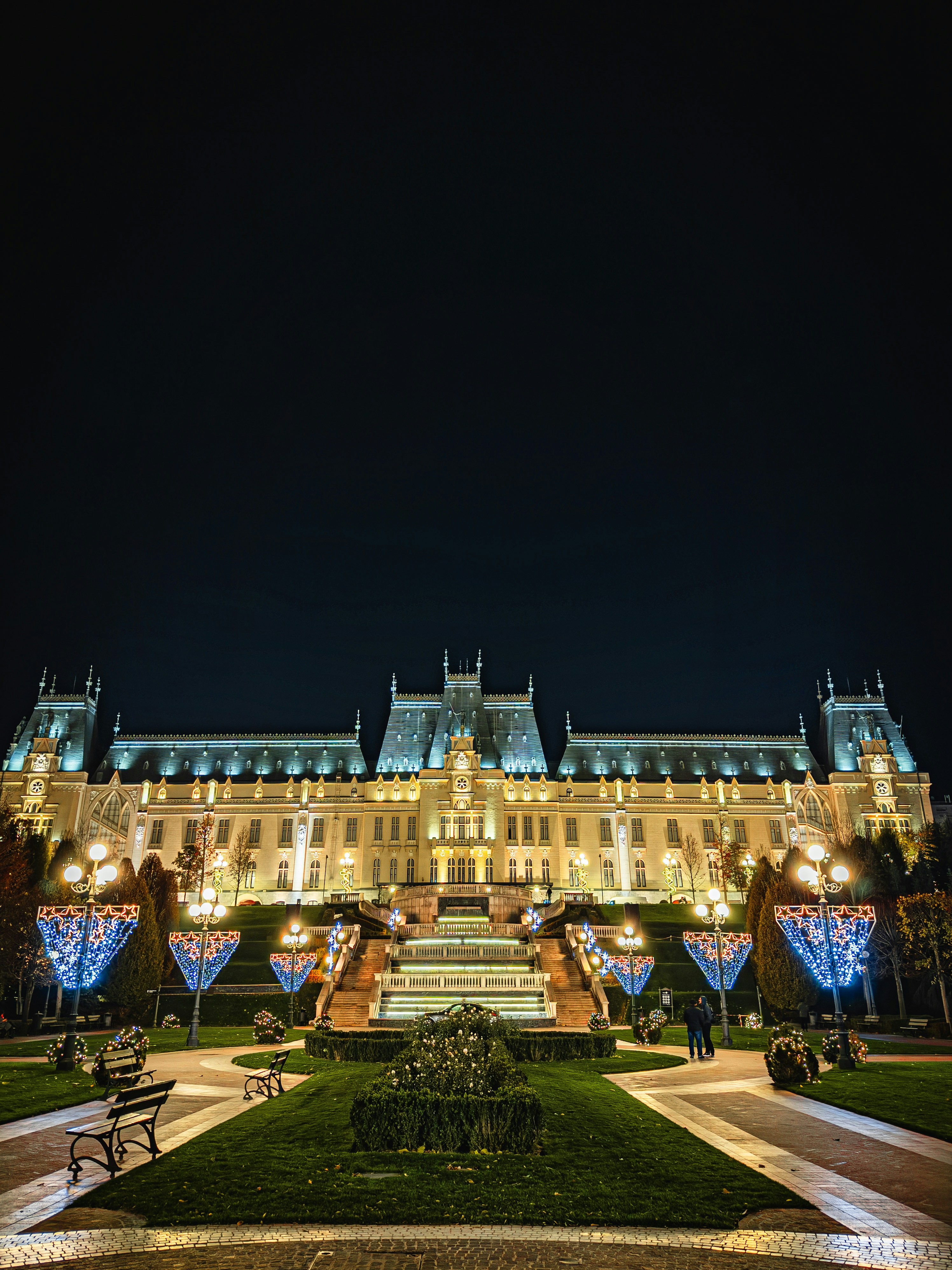 Historic palace adorned with festive lights, surrounded by manicured gardens and decorative trees. Night sky enhances the architectural details.