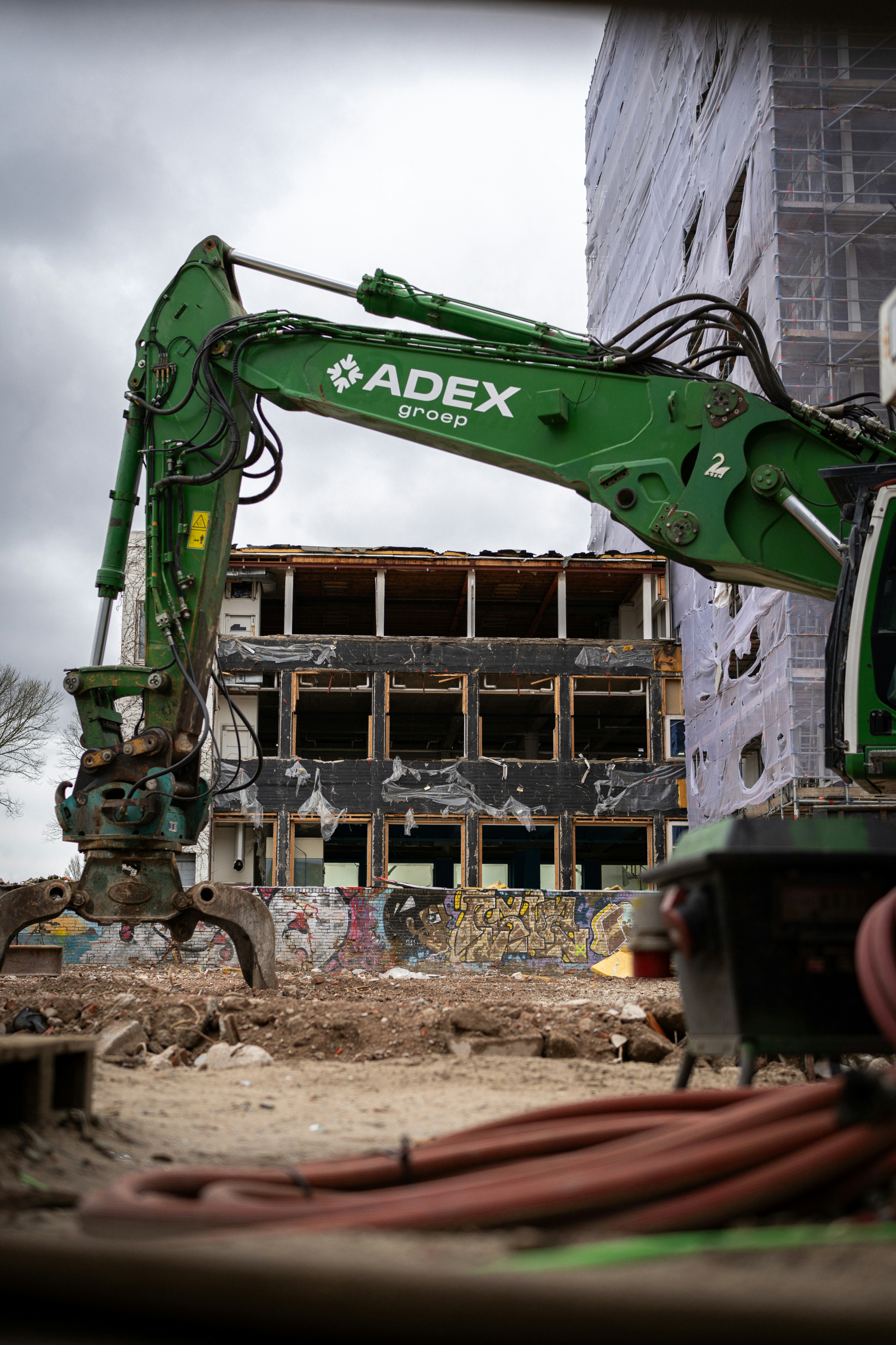 Green excavator arm frames a weathered construction site beyond a partially demolished building