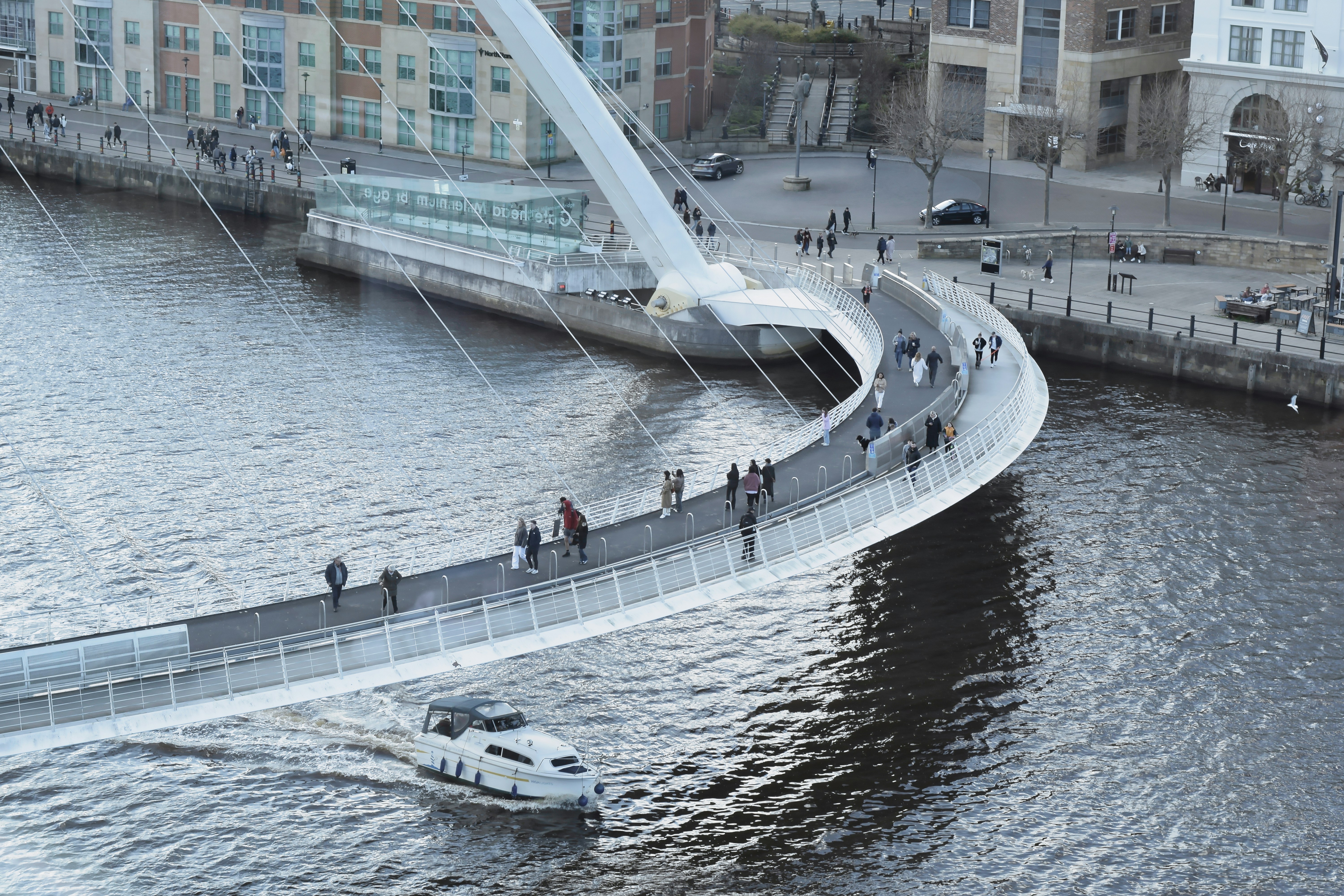 Curved pedestrian bridge arches over a calm river with a boat passing beneath and city buildings in the background.