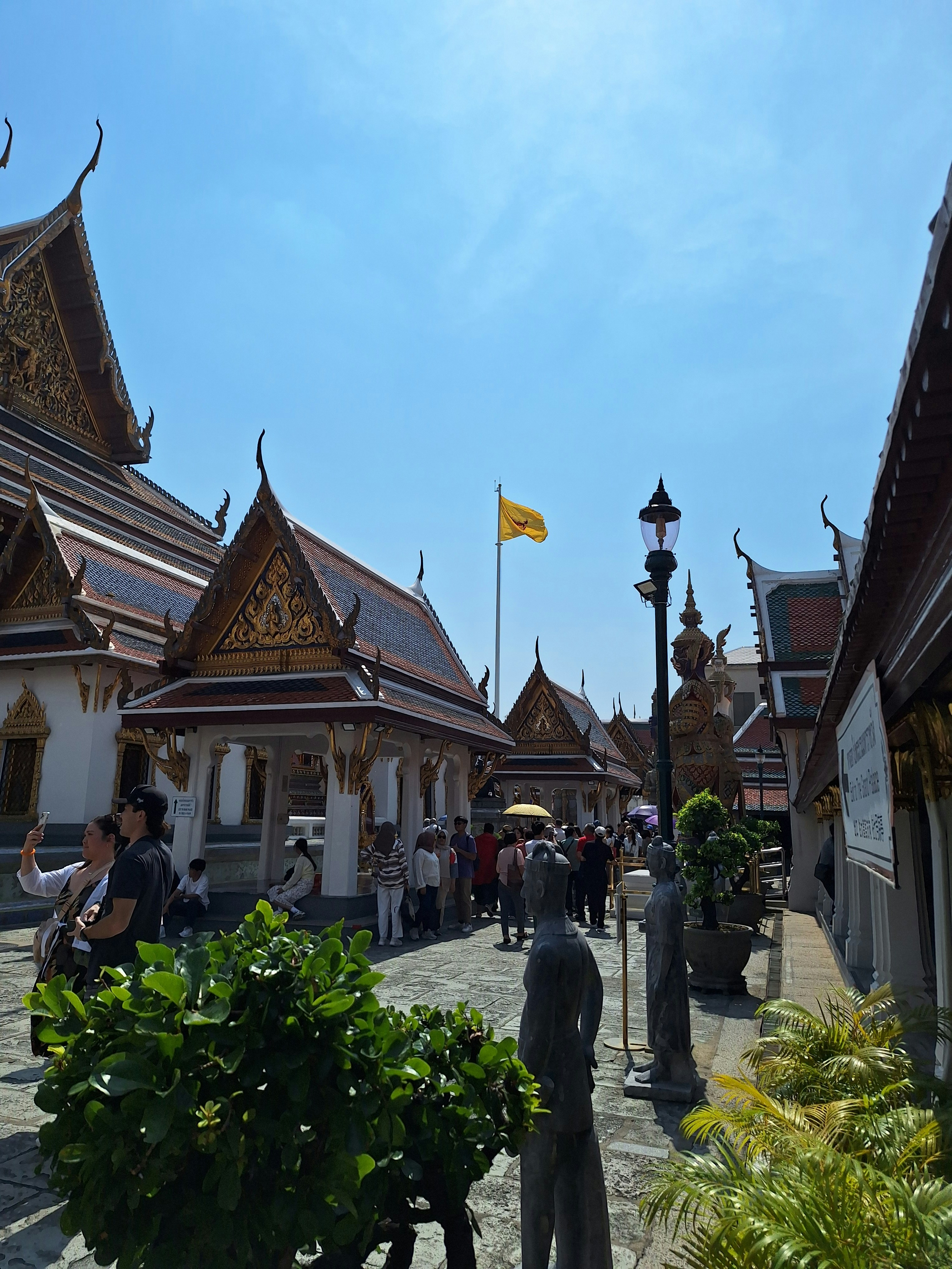 Sunlit temple complex with ornate, multi-tiered roofs and gold detailing; visitors stroll through a courtyard framed by stone statues and lush plants.