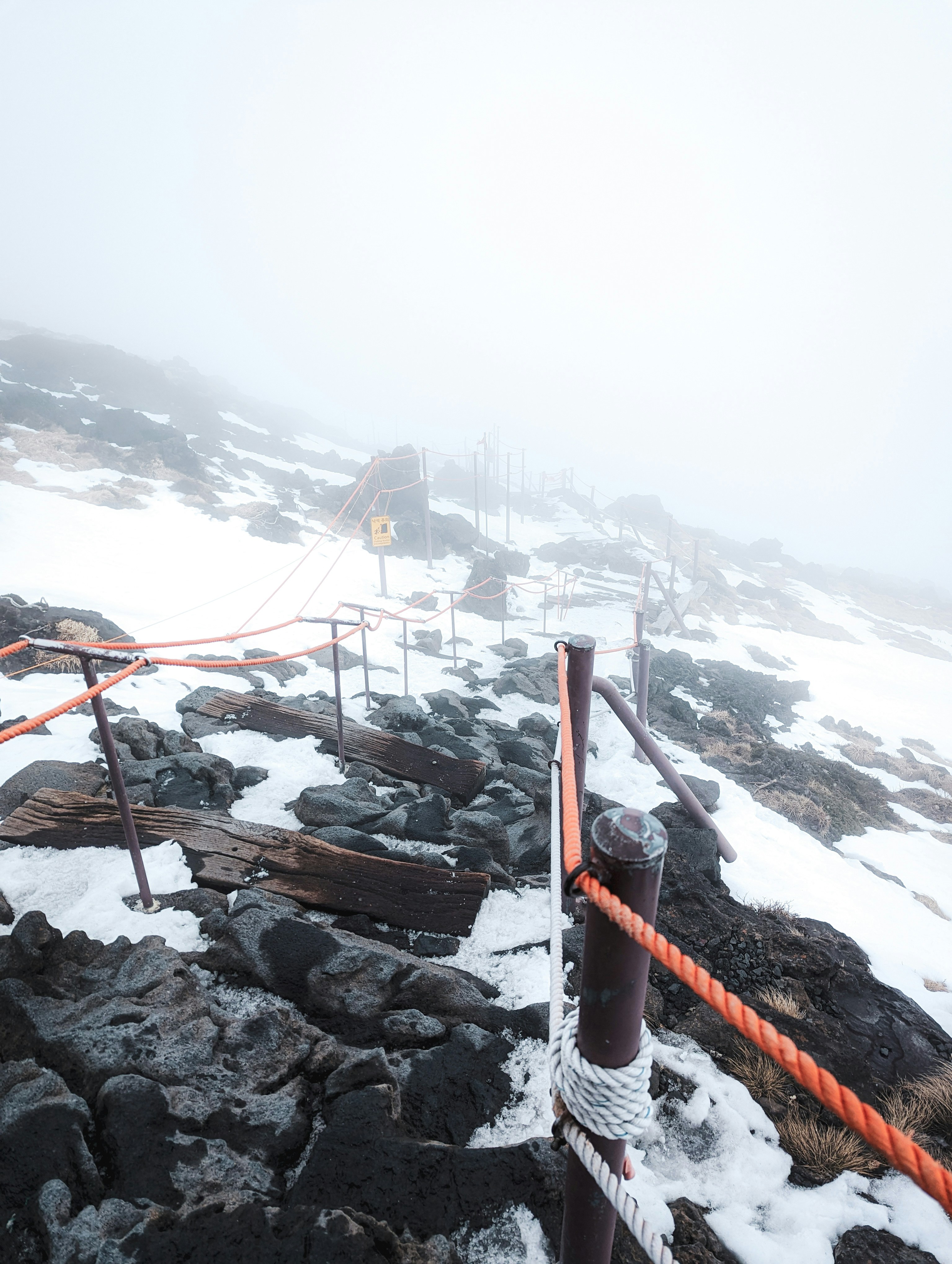 Rope-lined trail climbs a snow-dusted ridge into dense fog, with weathered posts and orange cords guiding the way. The scene emphasizes a rugged, windswept ascent into ethereal mist.