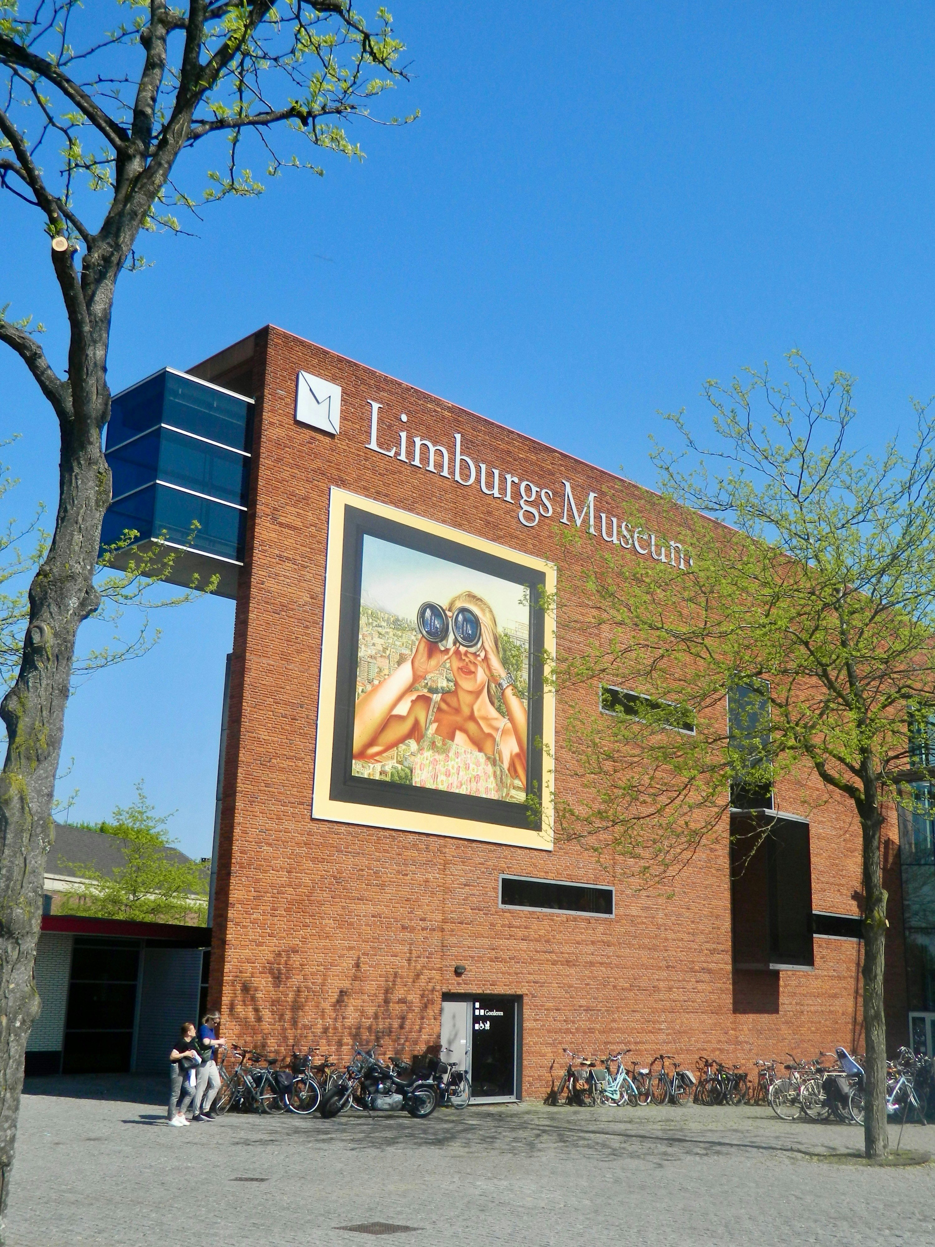 Brick building of Limburgs Museum with large mural depicting a person with binoculars, surrounded by bicycles under a clear blue sky.