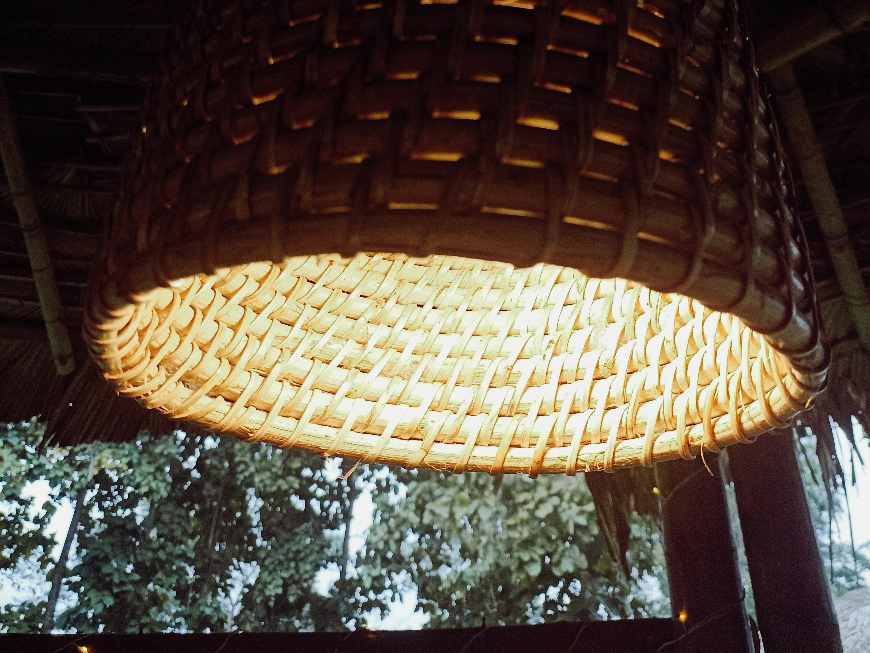 Close-up of a woven wicker lantern hanging from a thatched roof with a leafy outdoor backdrop.