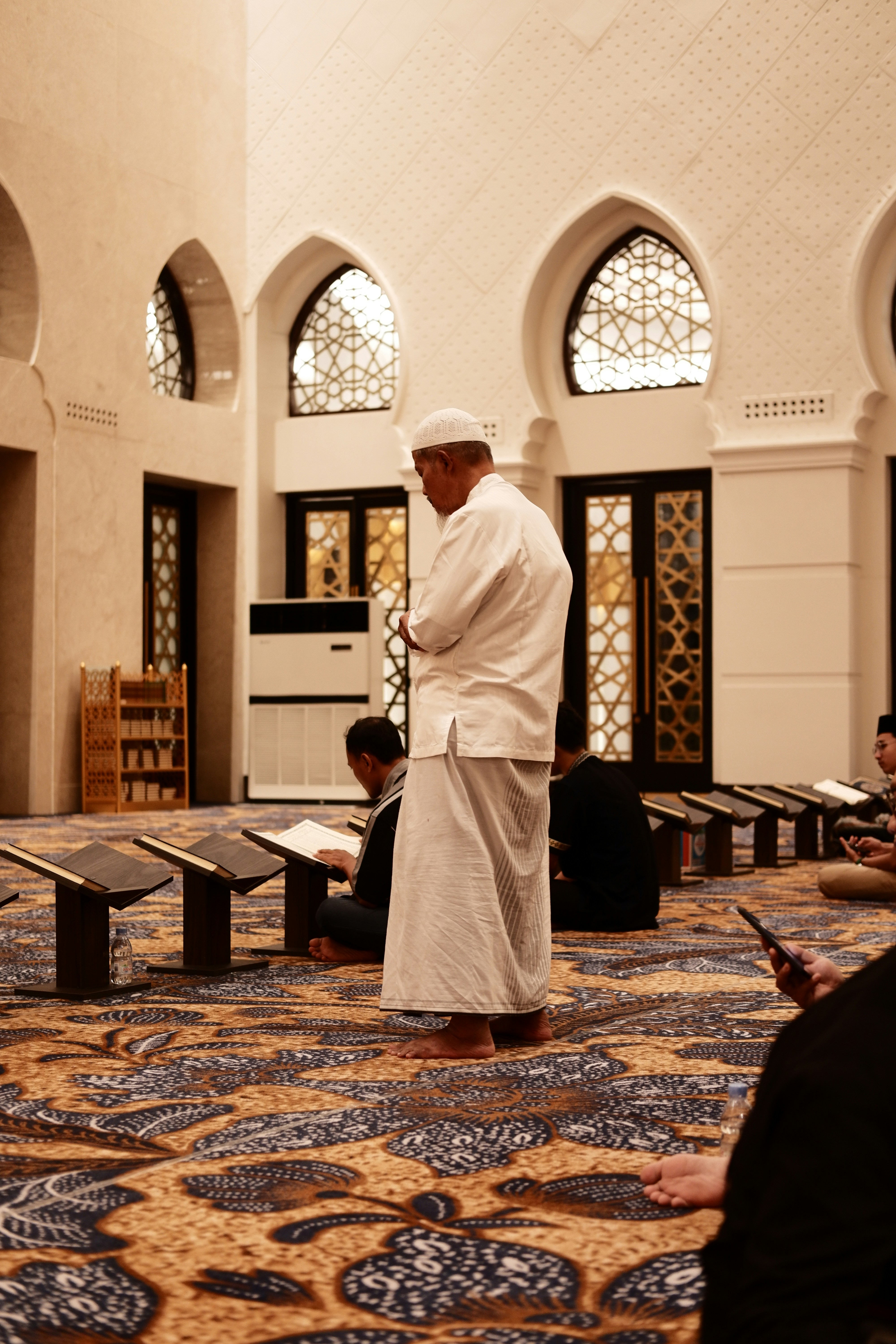 A photograph capturing a white-robed worshipper standing in prayer among others on prayer mats inside a grand hall with arched lattice windows.