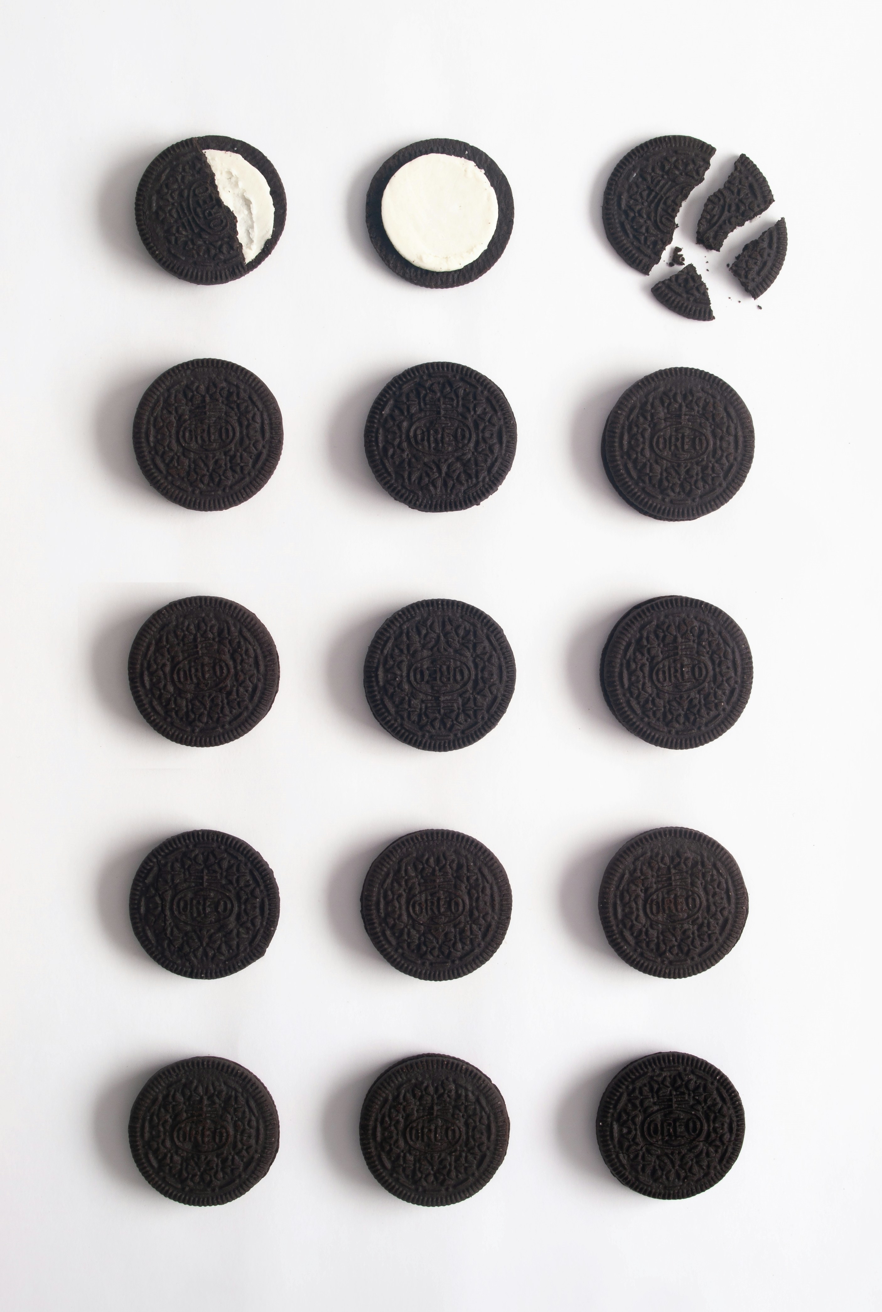 A tidy 3x5 grid of dark sandwich cookies on a white background, with the top-right cookie cracked open to reveal the cream.