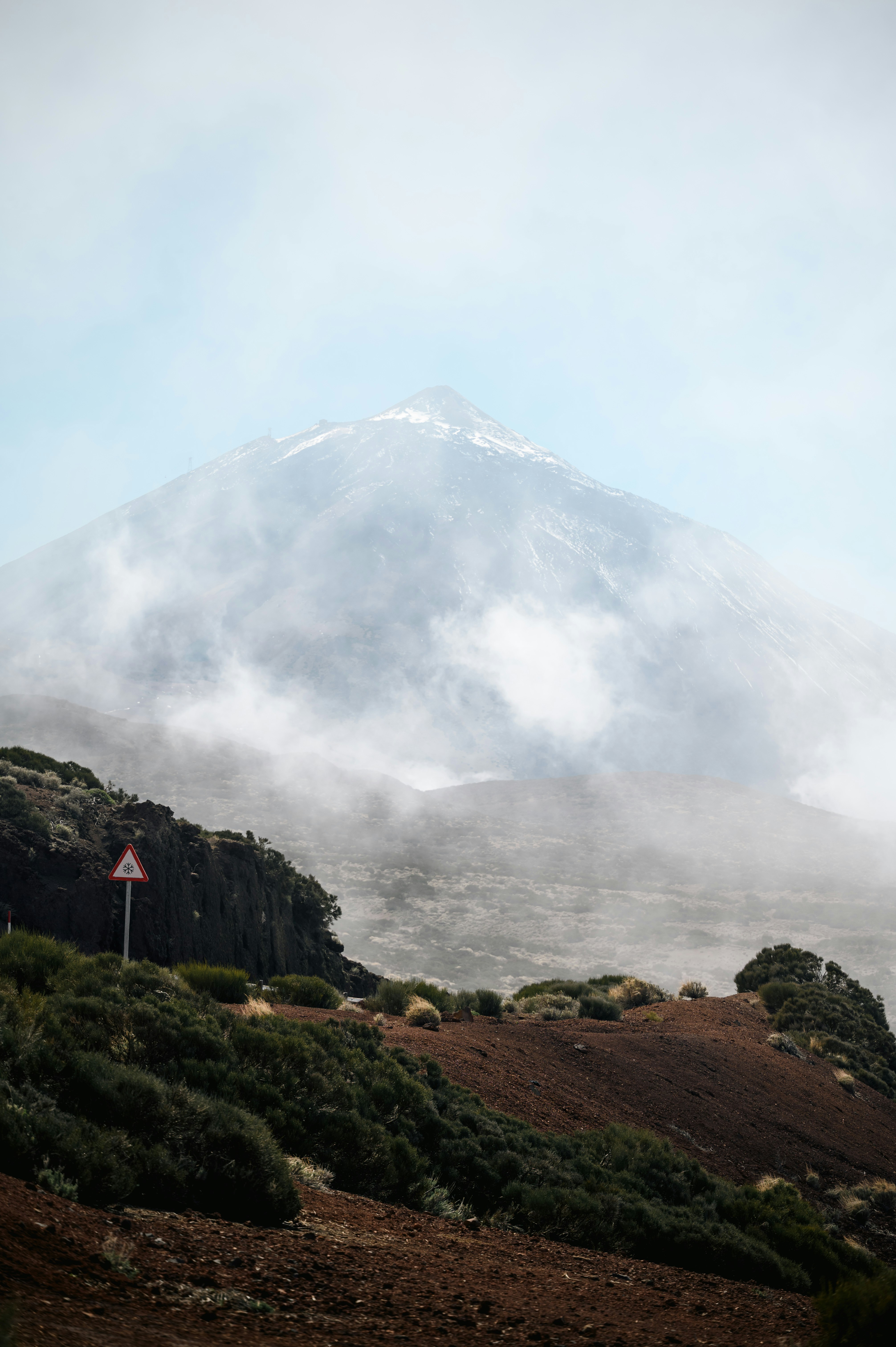 Snow-capped mountain emerging through a veil of mist, surrounded by rugged terrain and sparse vegetation.