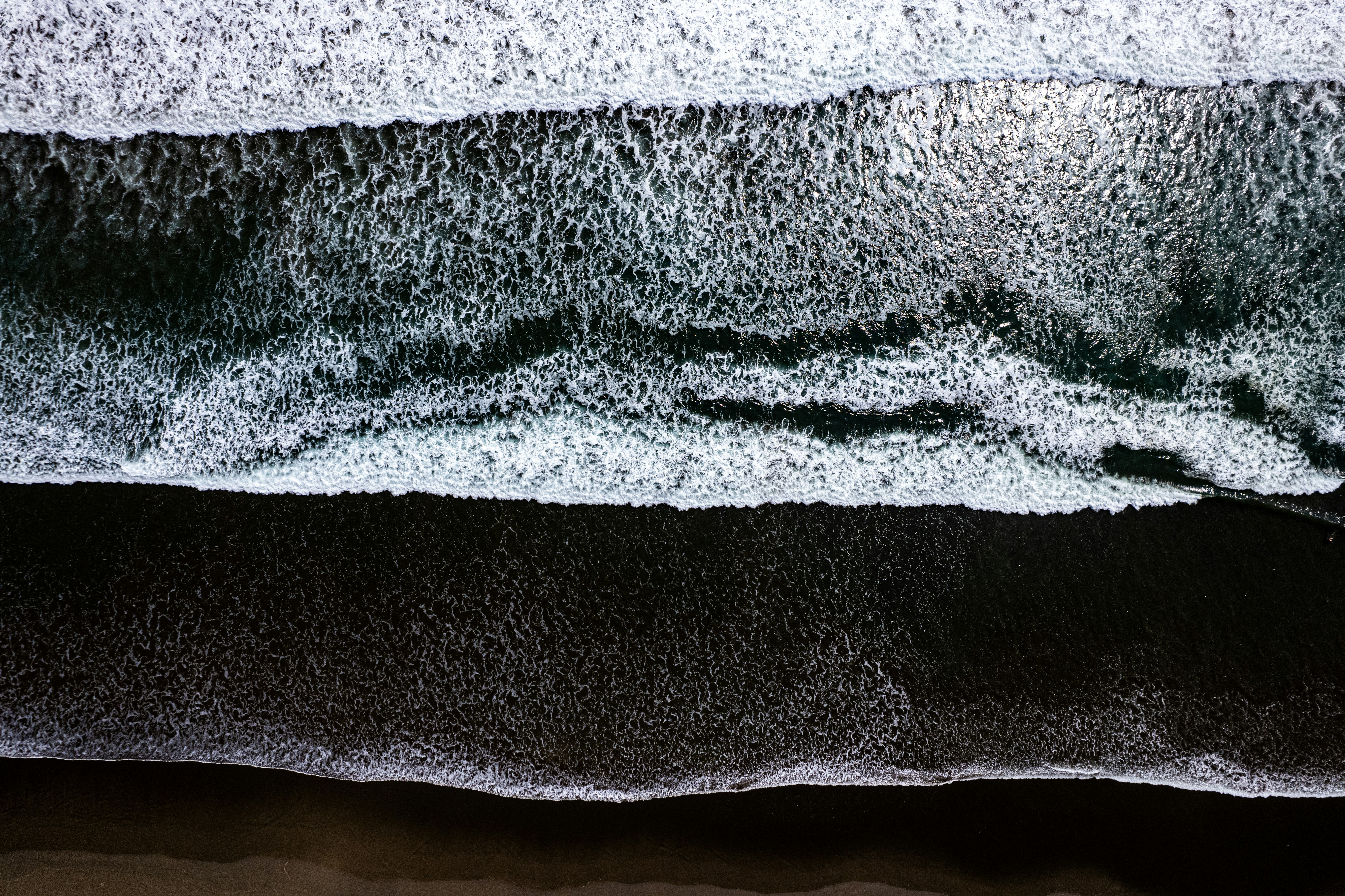 Aerial view of dark volcanic sand meeting white frothy waves with intricate ocean patterns.