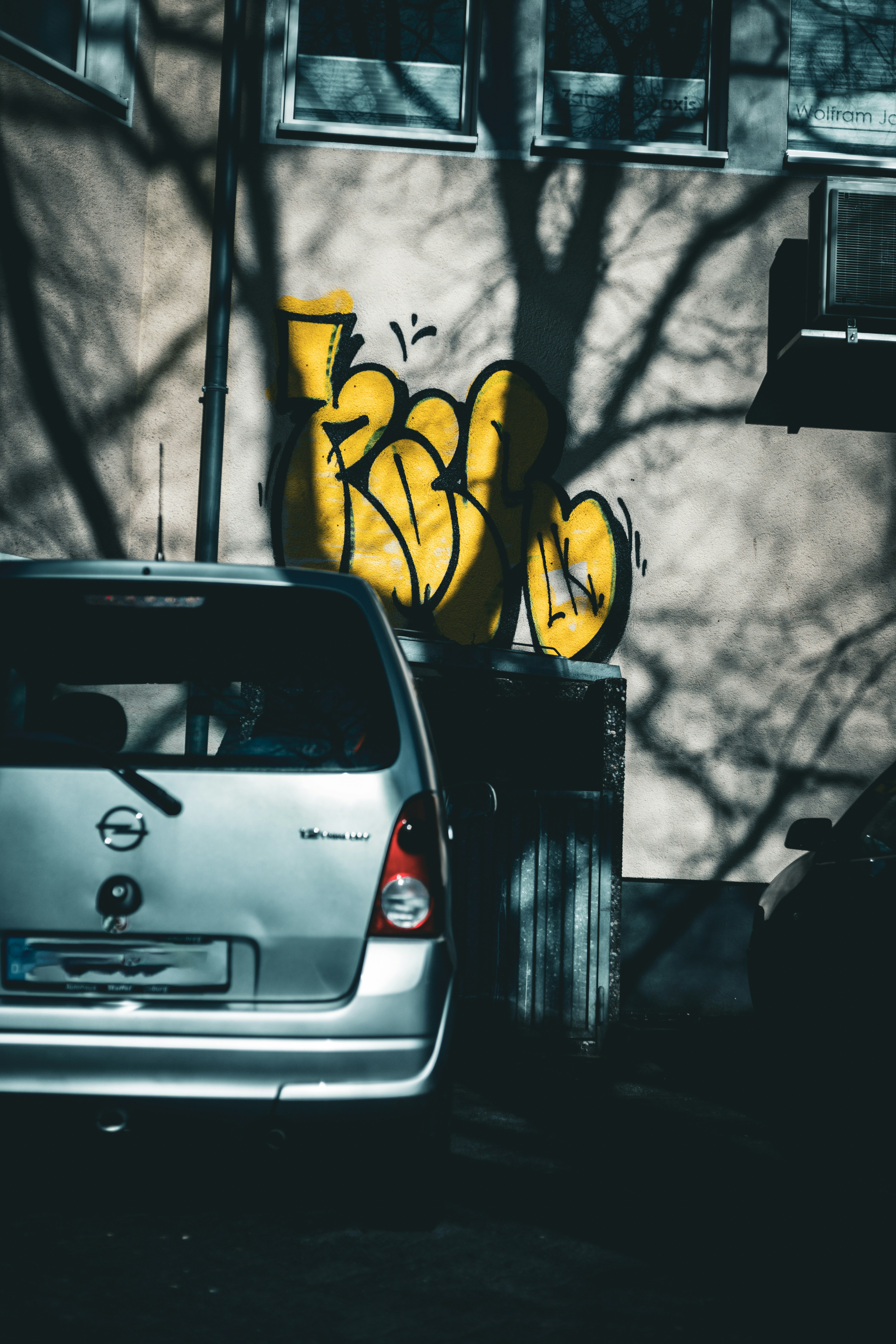 Yellow graffiti on a building wall with tree shadows and a parked car in the foreground.