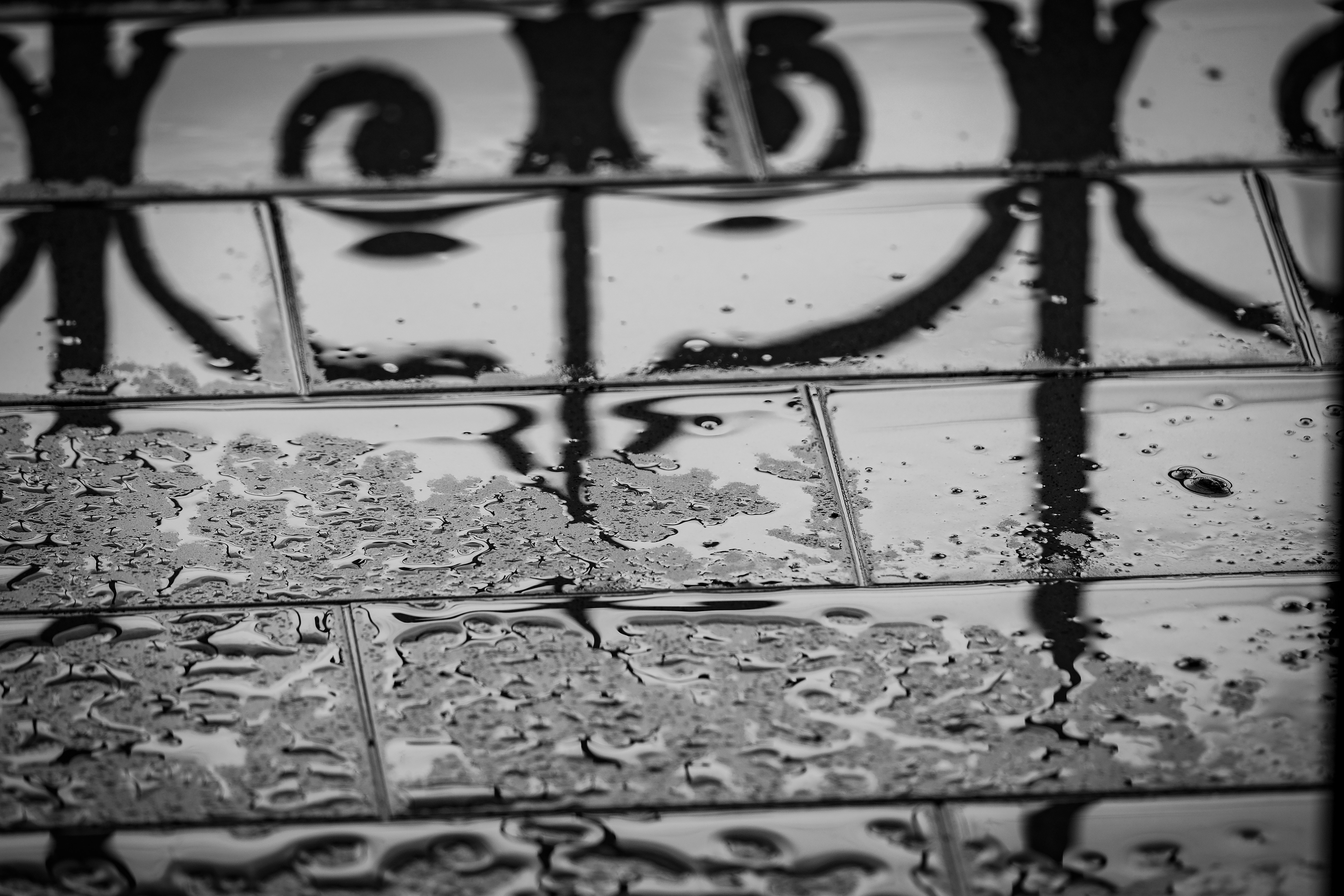 Black-and-white close-up of a wet tiled pavement with raindrop textures and a blurred iron railing reflection, highlighting surface texture and abstract pattern. This is a photographic still rather than an illustration or logo.