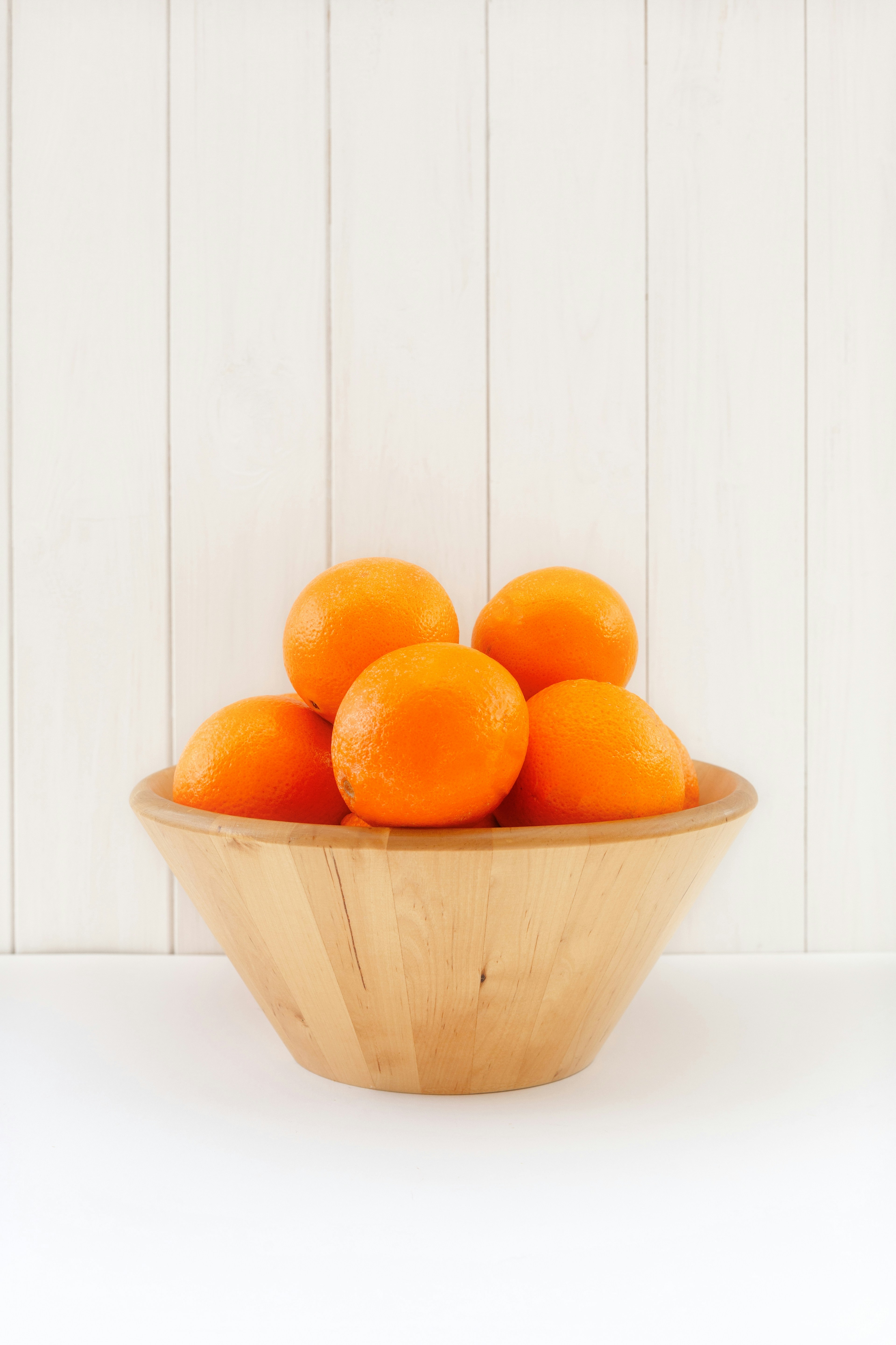 A wooden bowl filled with vibrant oranges, set against a clean white backdrop. The arrangement highlights the natural beauty and freshness of the fruit.