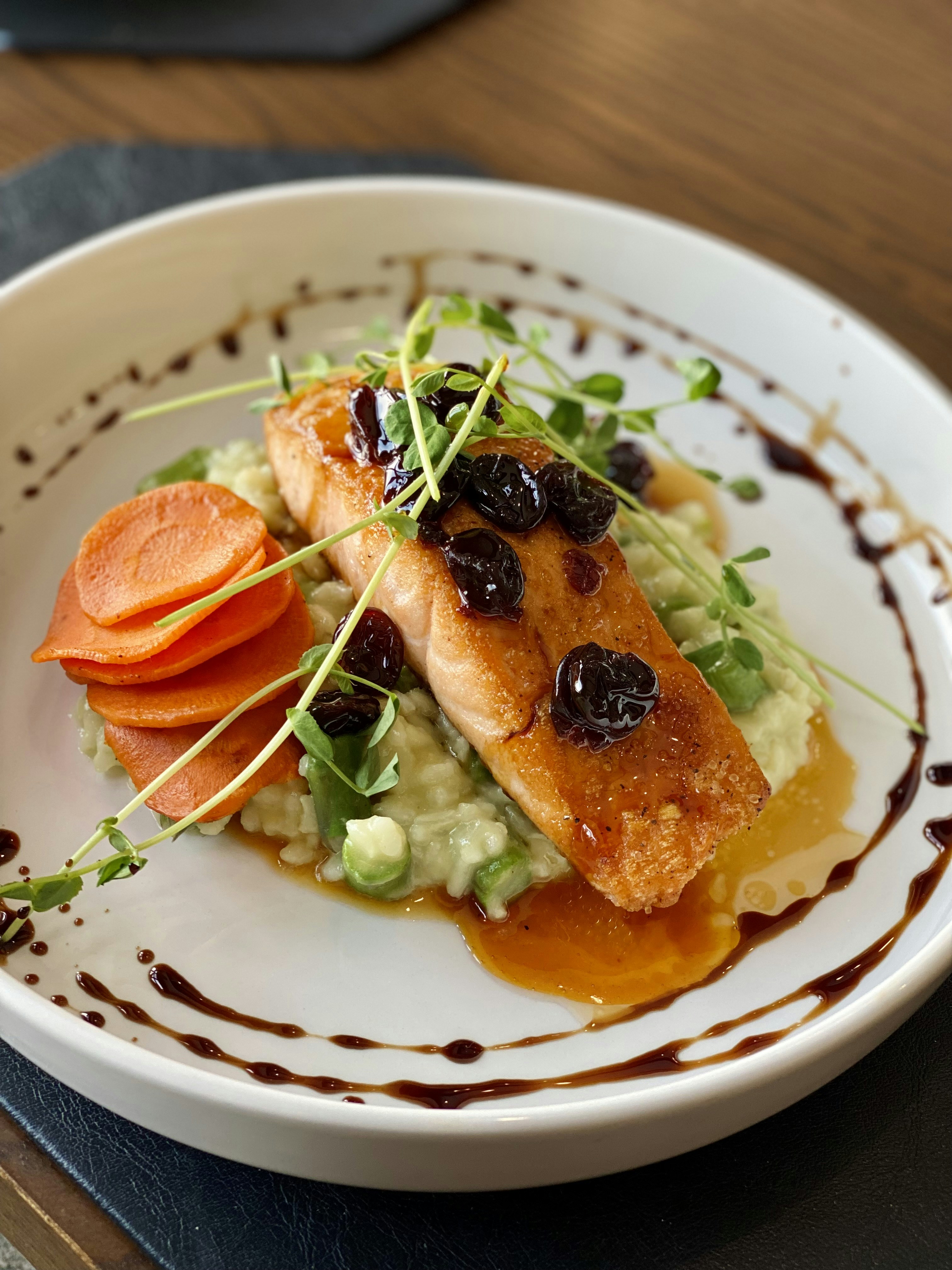 Grilled salmon fillet garnished with cherries and microgreens, served atop a creamy risotto with vibrant carrot slices. 