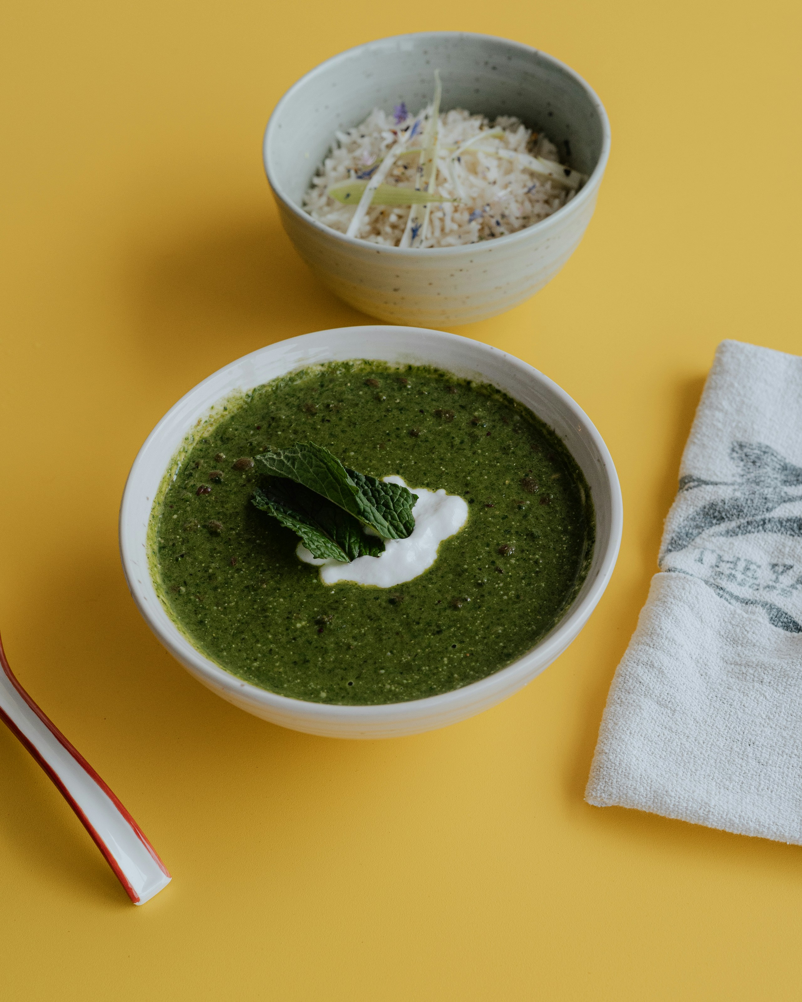Bowl of vibrant green soup garnished with mint leaves and a dollop of cream, accompanied by a bowl of rice and a folded napkin on a bright yellow background.