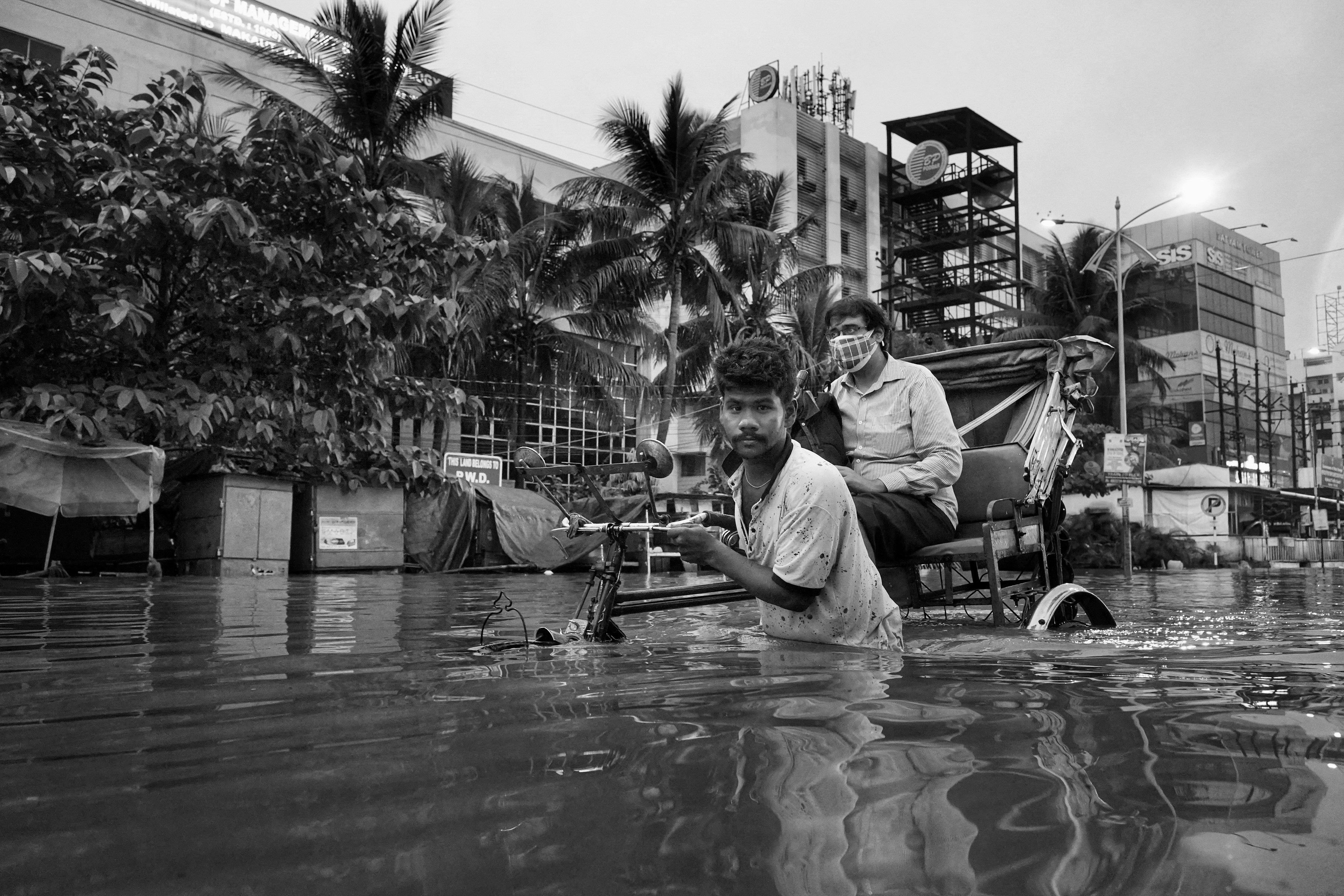 Man pushing a rickshaw through a flooded street amidst urban buildings and palm trees.