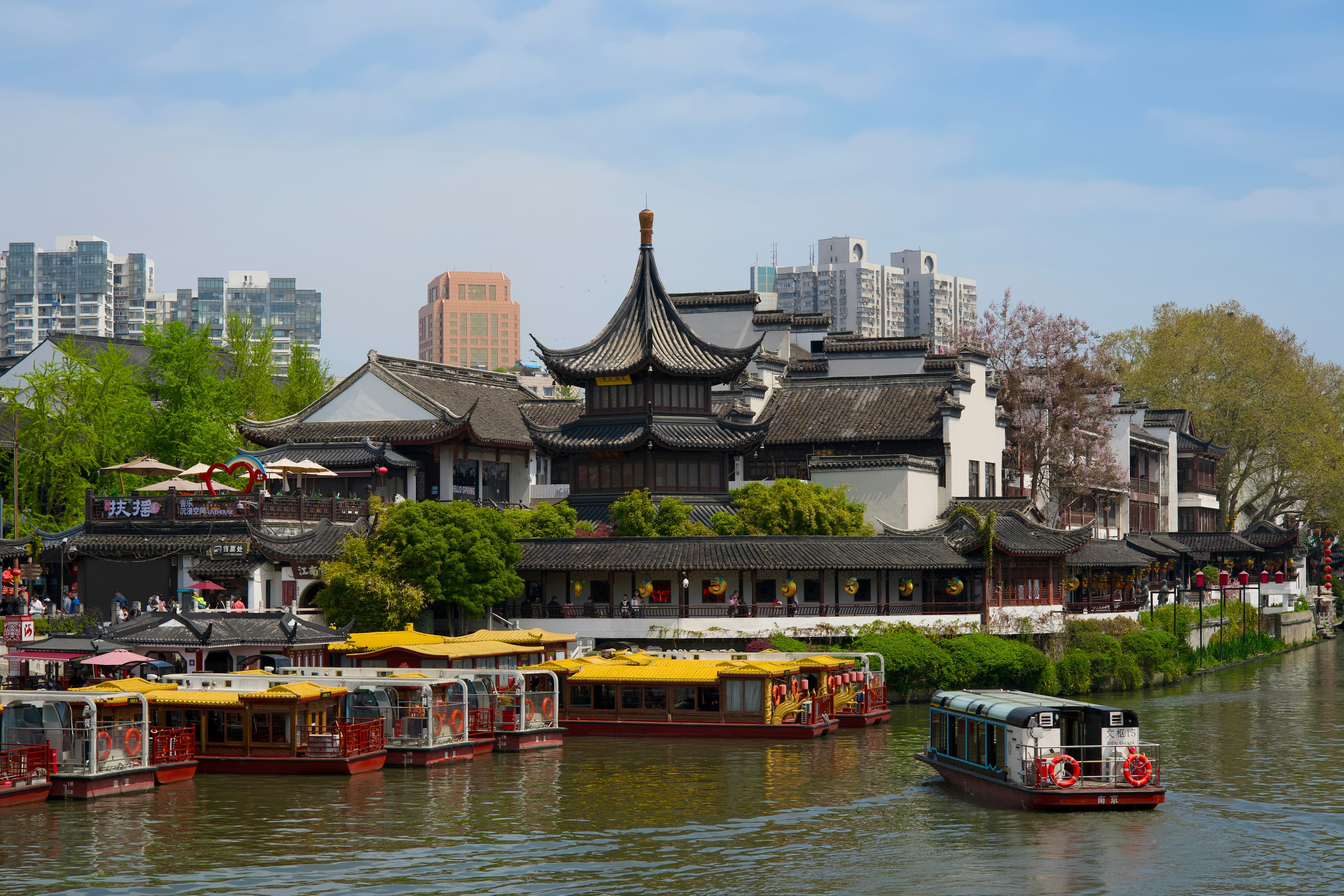 Traditional Chinese buildings along a river with boats under a clear blue sky.