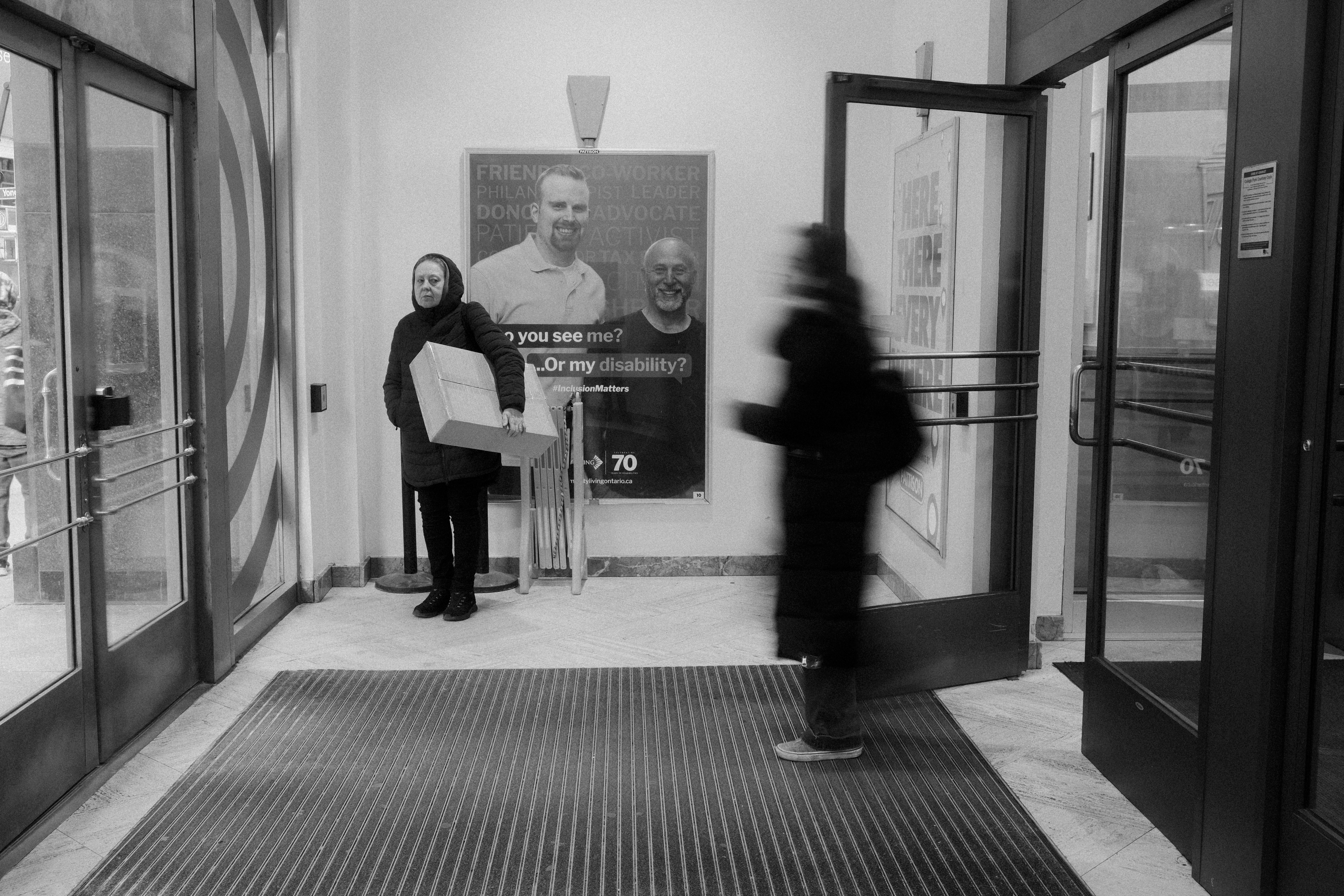 Black and white scene of a woman holding a box near a revolving door in a busy entranceway.