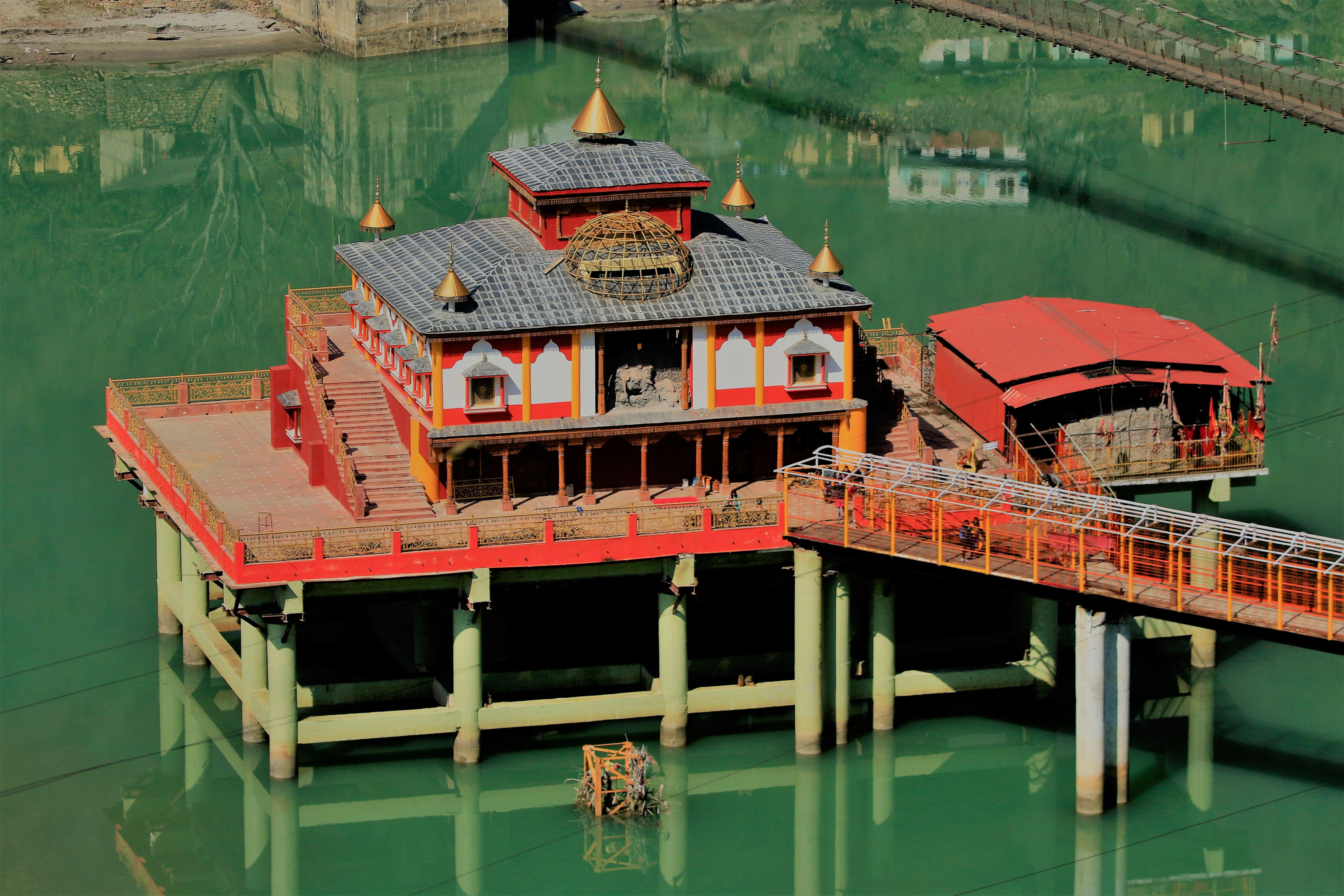 Vibrant temple structure elevated above calm green water with a connecting walkway.