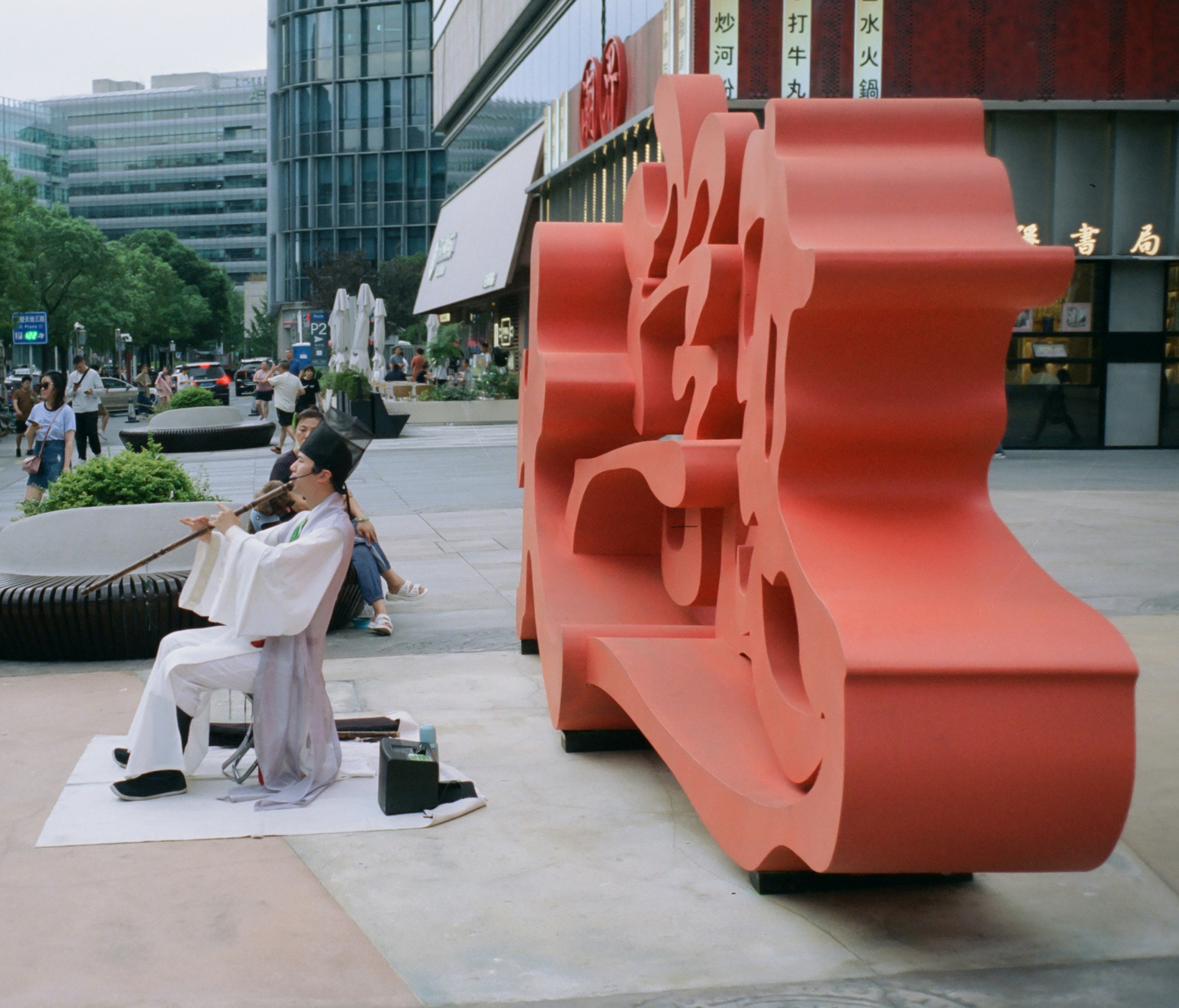 A street musician plays a flute beside a towering red abstract sculpture in a modern urban plaza.