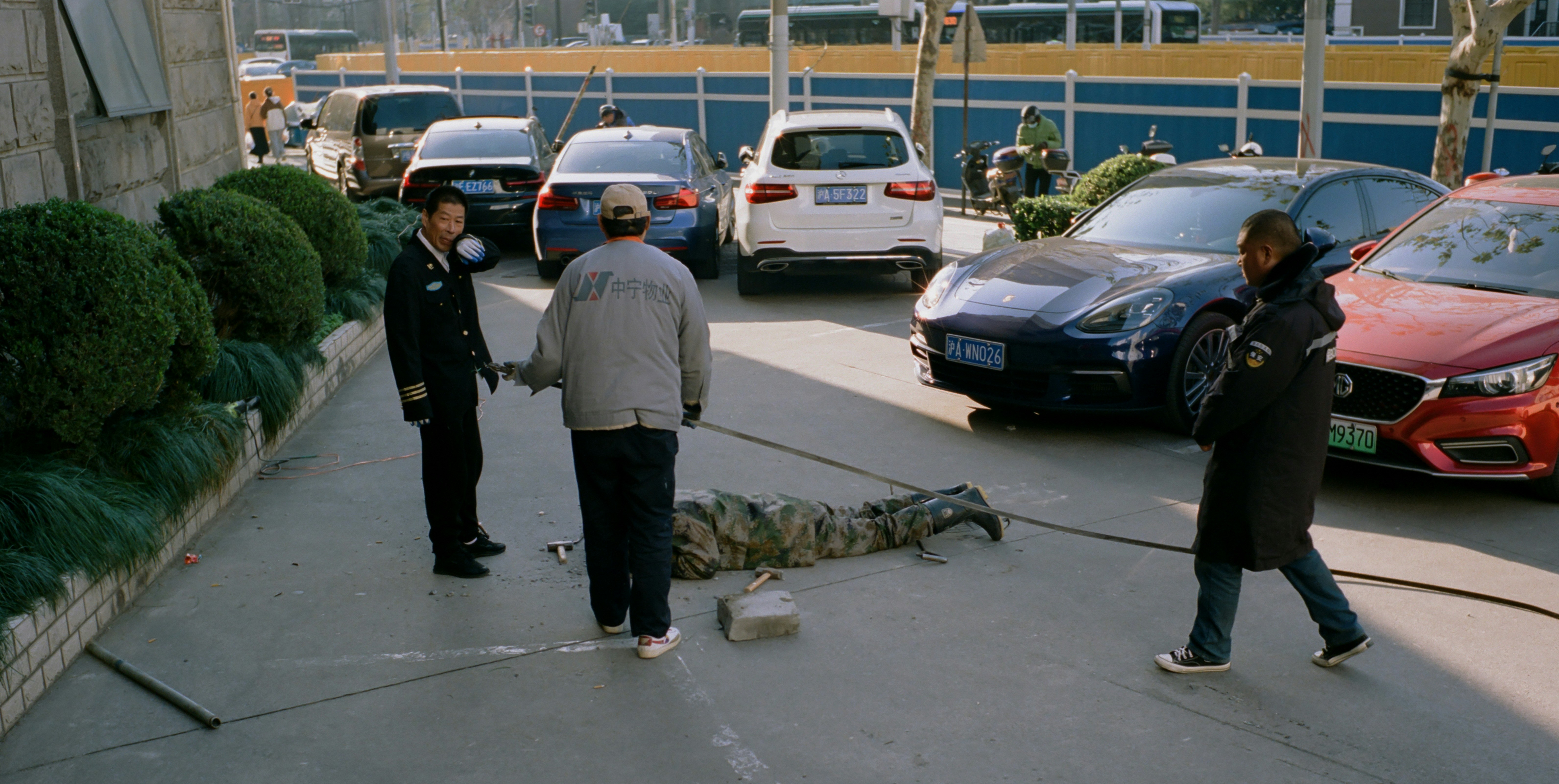 Three figures stand around a person on the ground in a parking lot, with several parked cars and a blue barrier in the background.