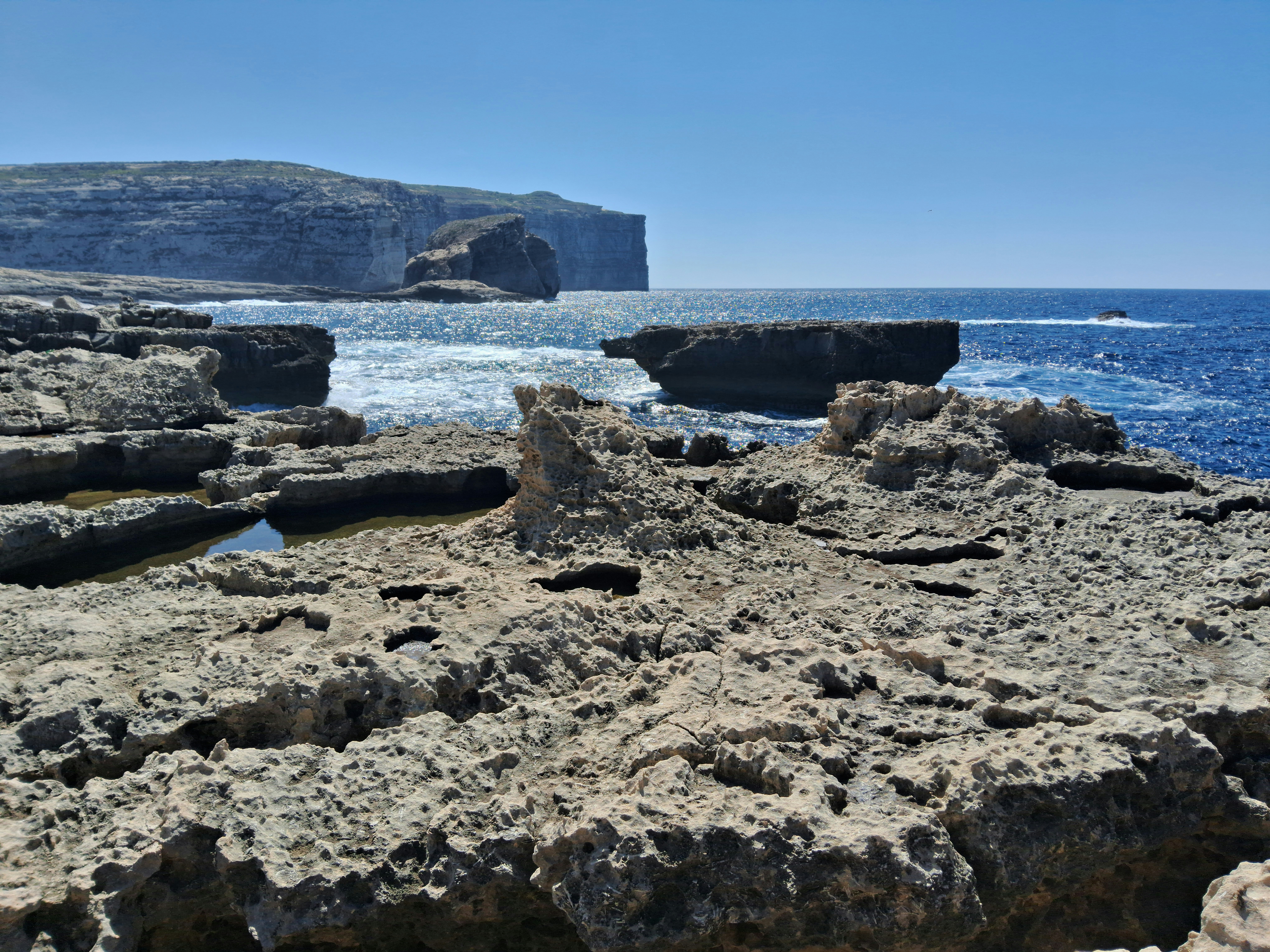 Rugged shoreline with weathered rock outcrops in the foreground and a distant cliff meeting a blue Atlantic under a clear sky. The texture and depth emphasize the coastal geology and expansive horizon.