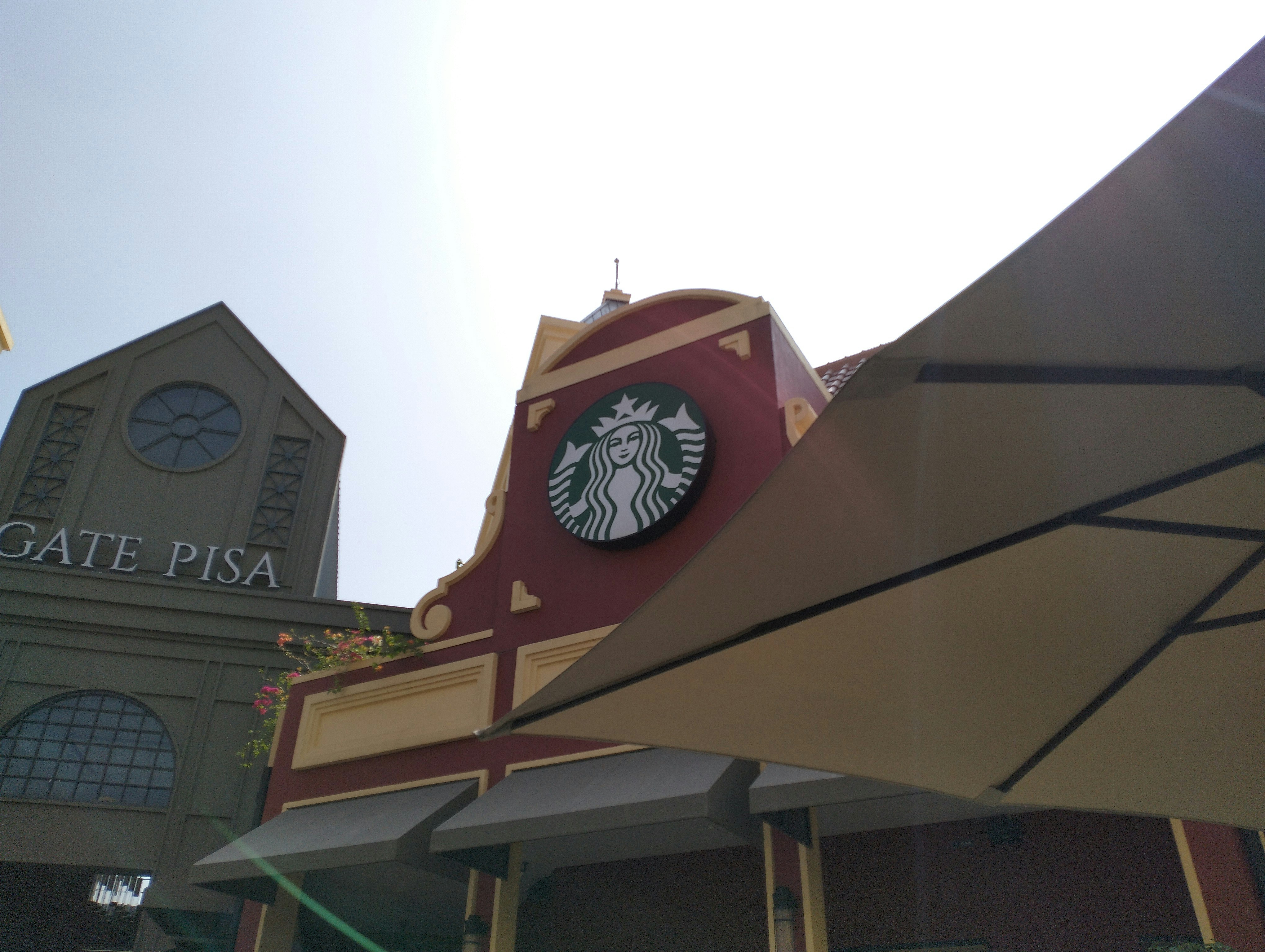 Sunlit cafe exterior featuring a prominent Starbucks logo on a red façade beside a large patio umbrella.
