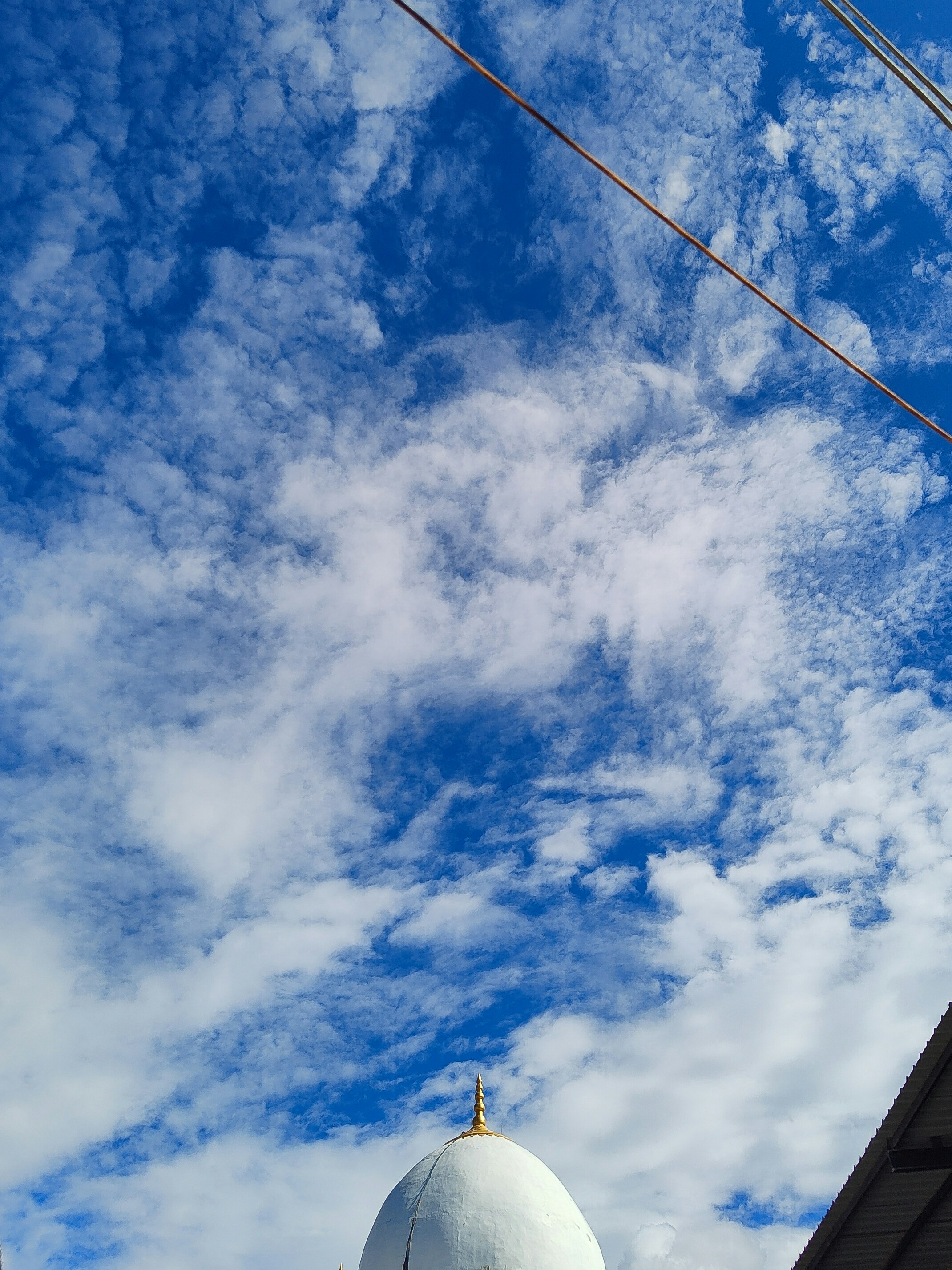 White temple dome with a gold finial sits against a blue sky sprinkled with wispy clouds, intersected by overhead cables.