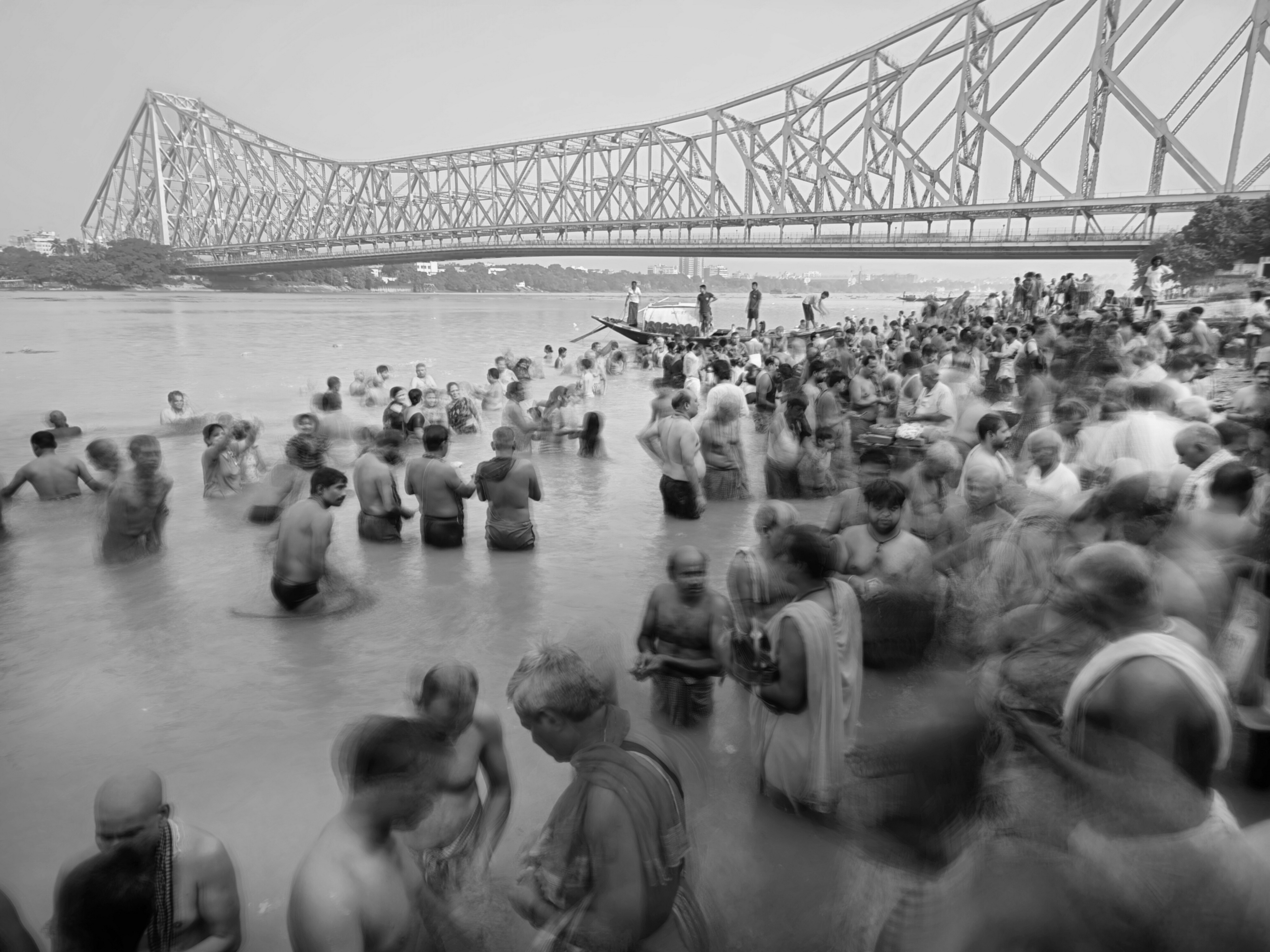 Monochrome photograph of a dense crowd bathing along the Hooghly River, with the Howrah Bridge spanning the background.