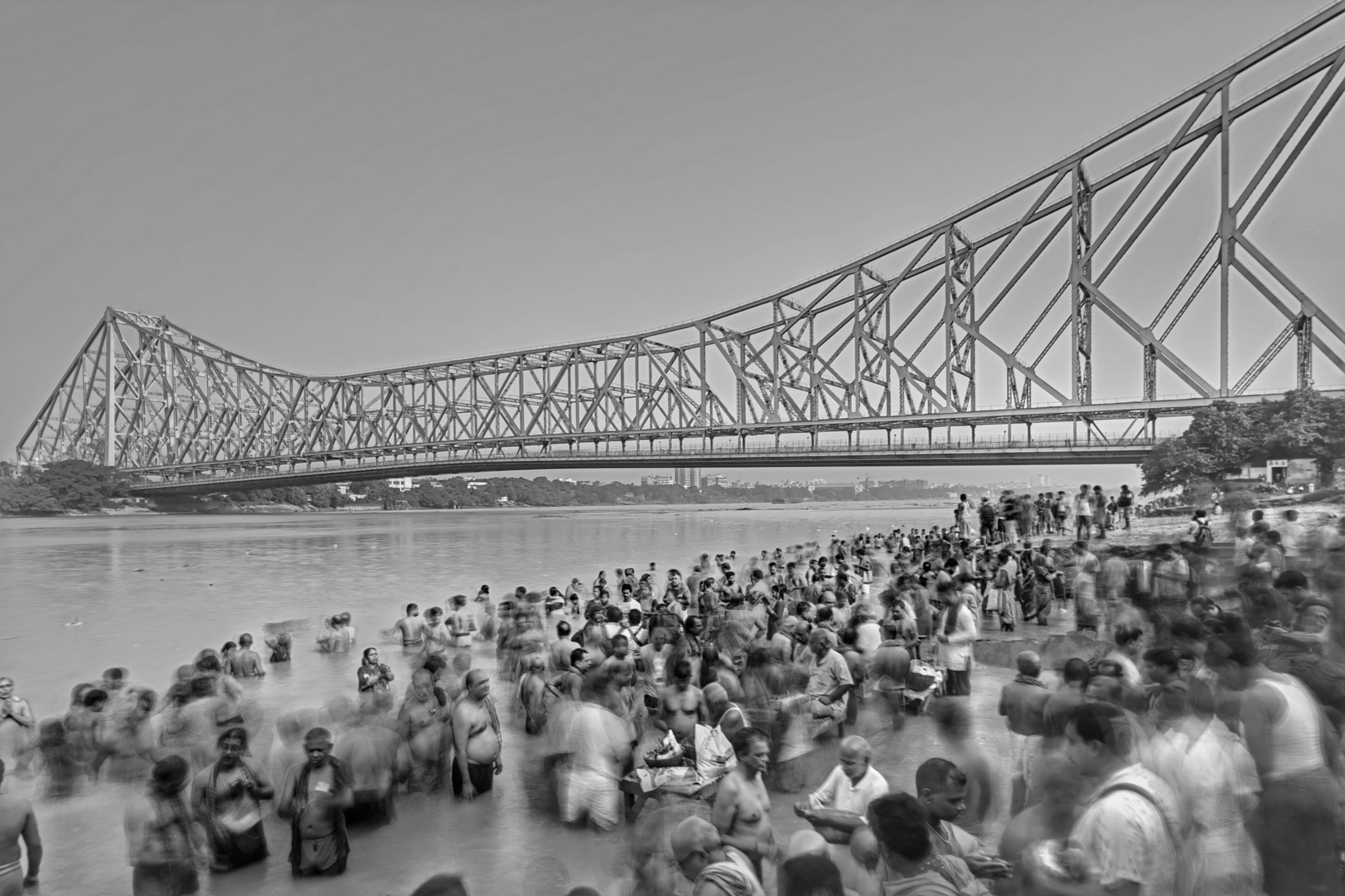 Monochrome photograph of a crowded beach with a long steel bridge spanning the water in the background. The dense crowd foreground emphasizes scale against the bridge.