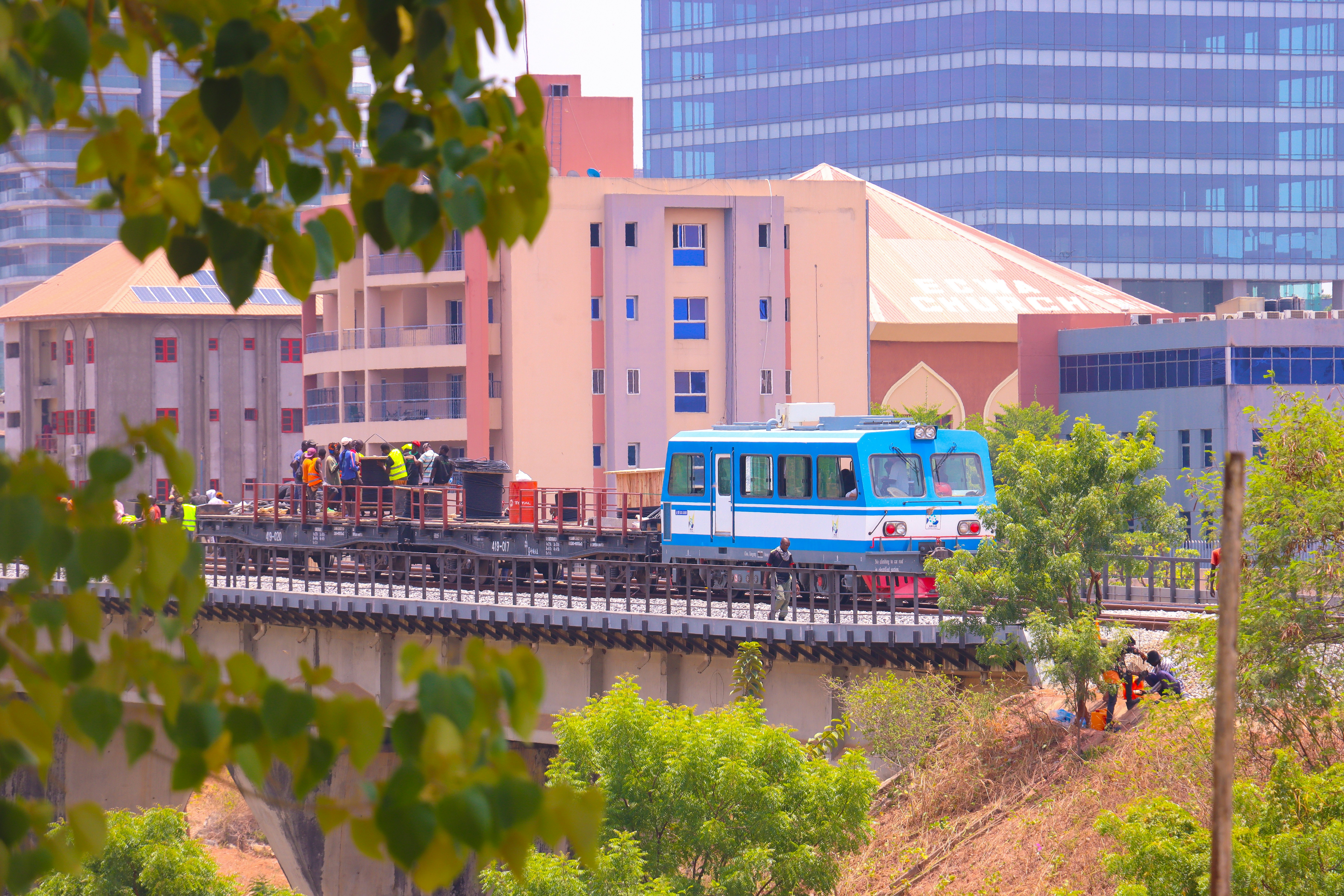 Blue train traversing a bridge amidst city buildings and greenery on a sunny day.