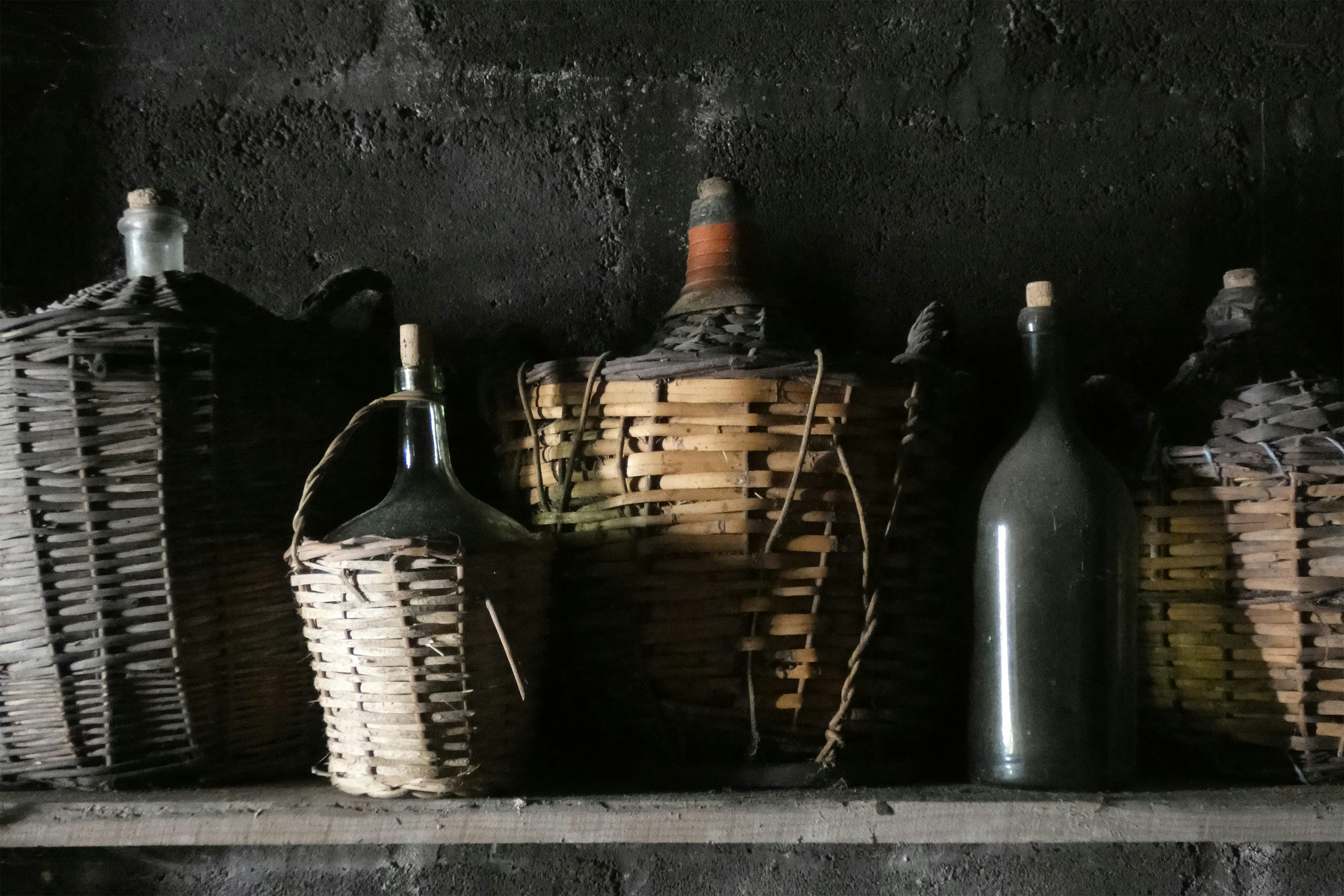 Dimly lit still-life photograph featuring weathered bottles and woven baskets resting on a dark shelf. The composition emphasizes texture and muted tones.