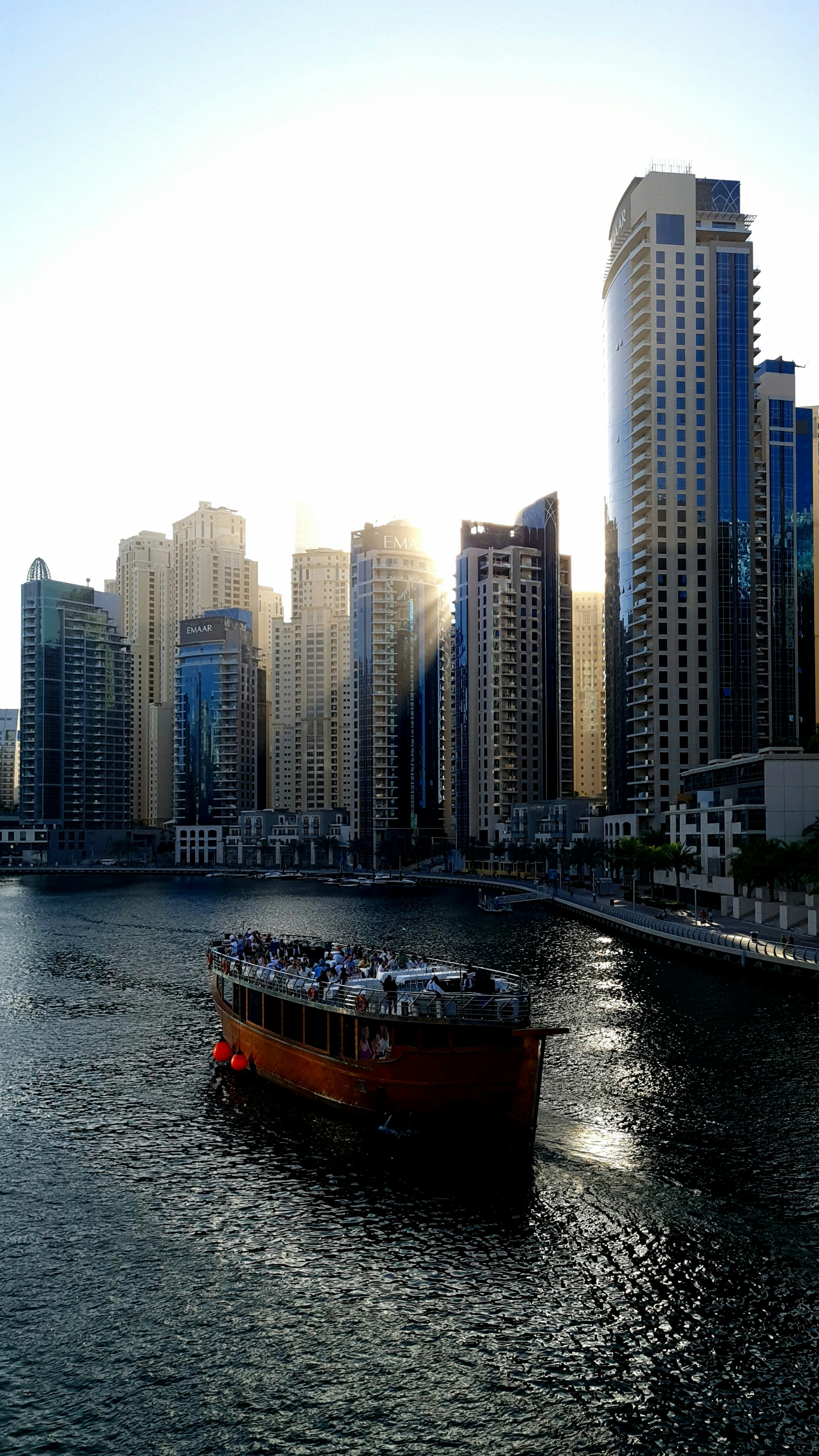 Photograph of a riverboat gliding through a marina flanked by towering glass skyscrapers at sunset.