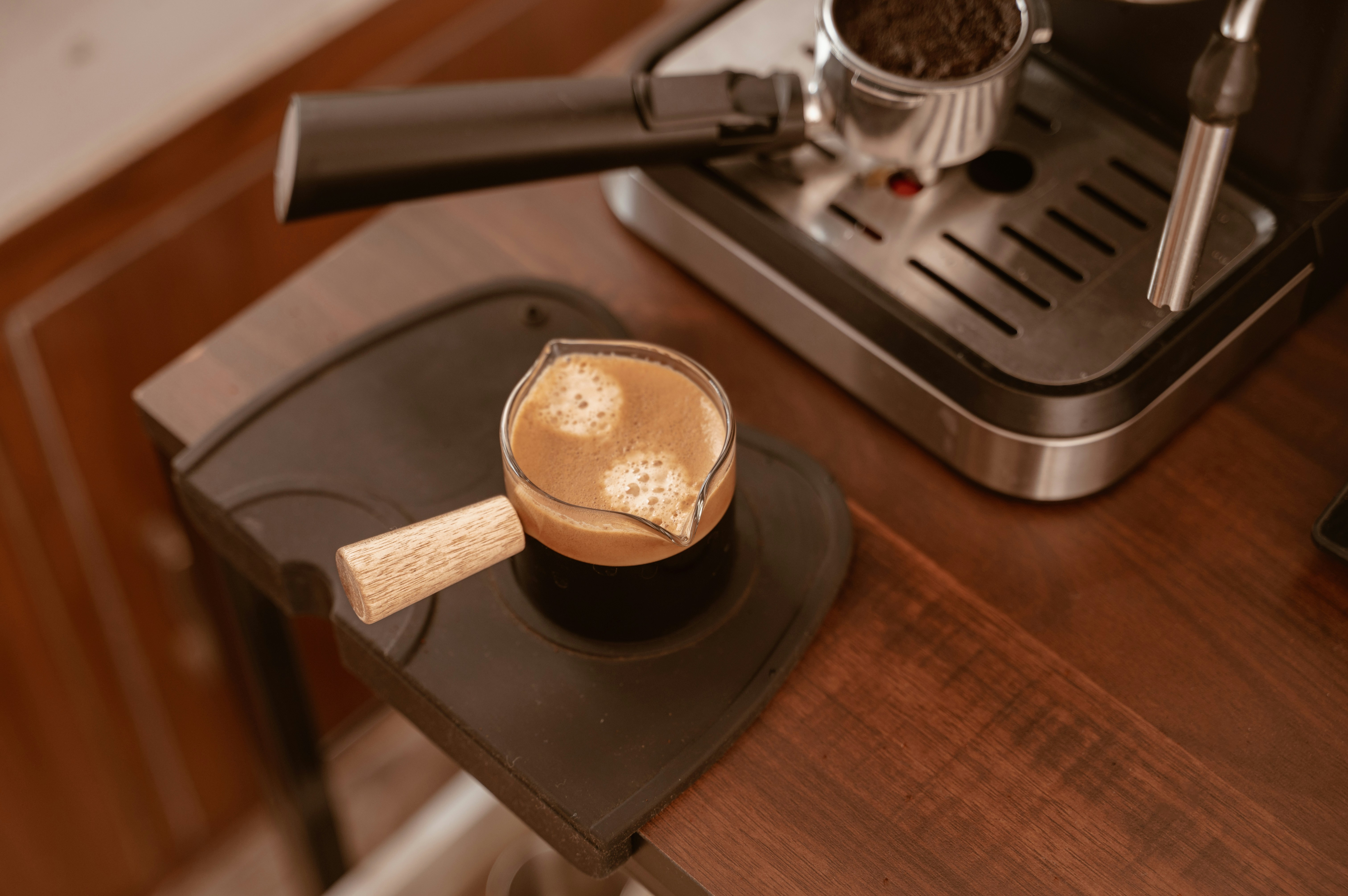 A freshly brewed espresso sits in a glass cup on a wooden countertop, with an espresso machine in the background. The scene captures the essence of coffee preparation.