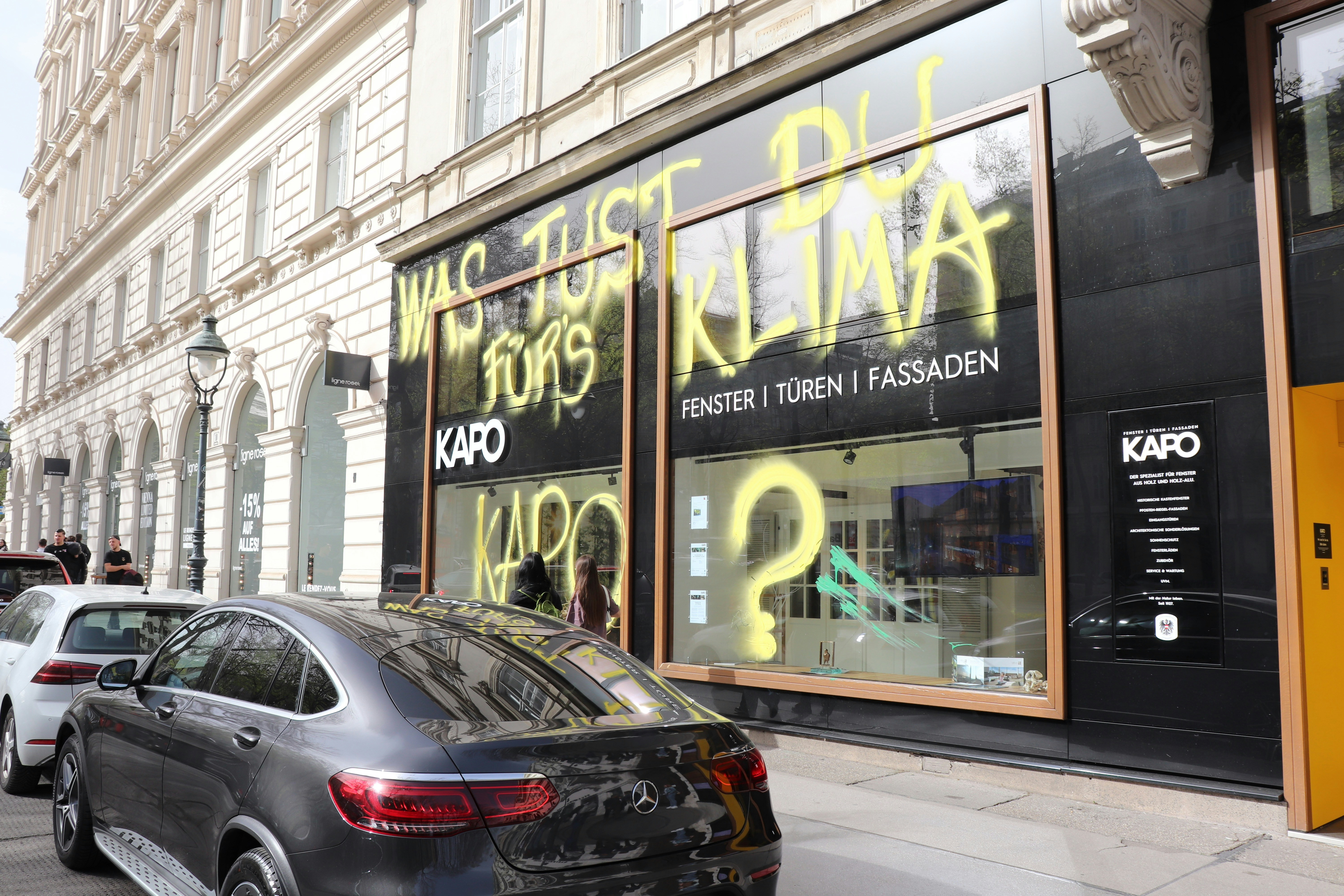 Bold graffiti in yellow letters covers a large glass storefront in an urban setting, with parked cars lining the street.
