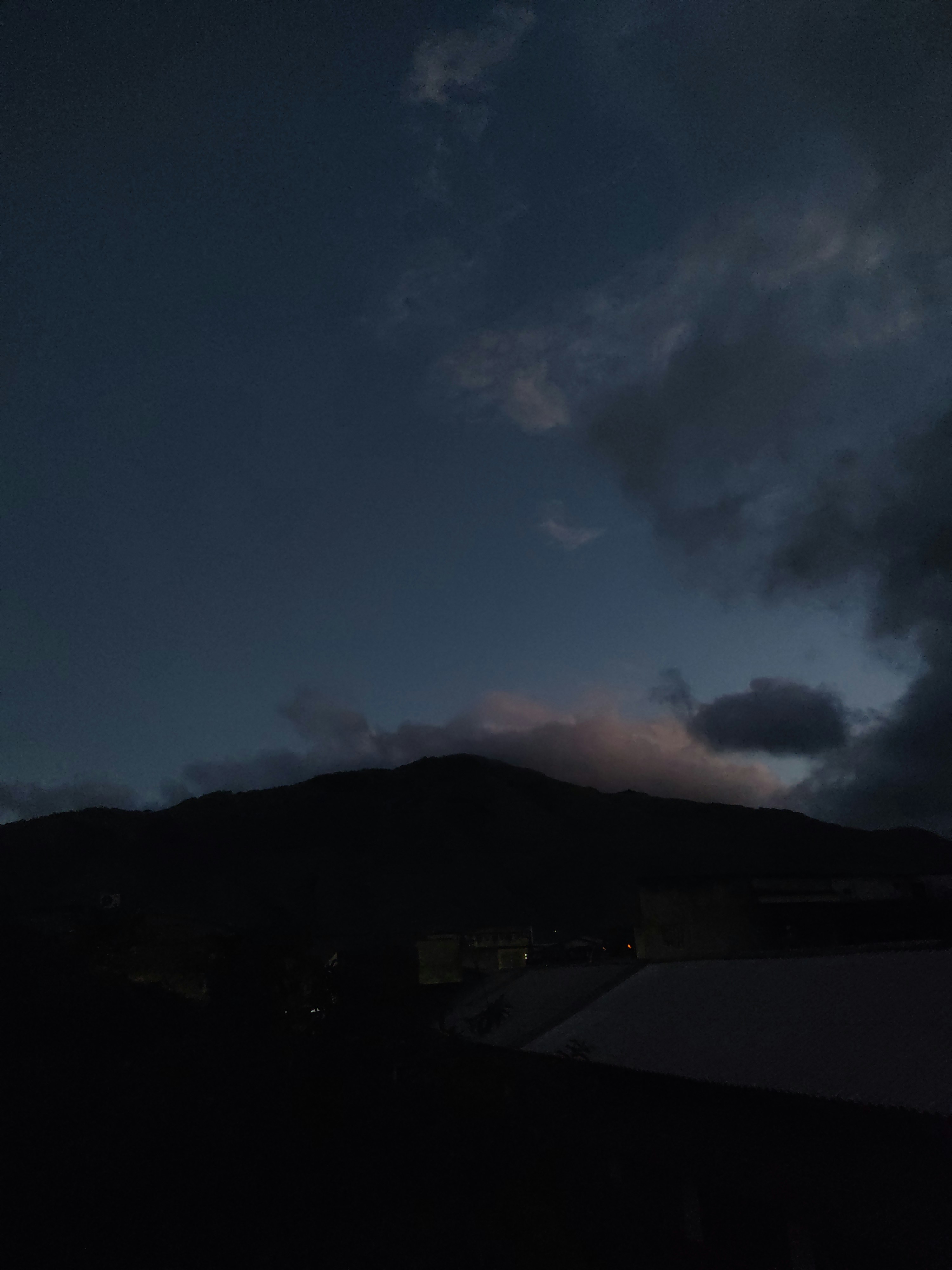 Dark landscape photograph of a silhouetted ridgeline under a dusky, blue sky with a pink-tinged cloud on the horizon. The frame emphasizes low light and atmospheric color at twilight.