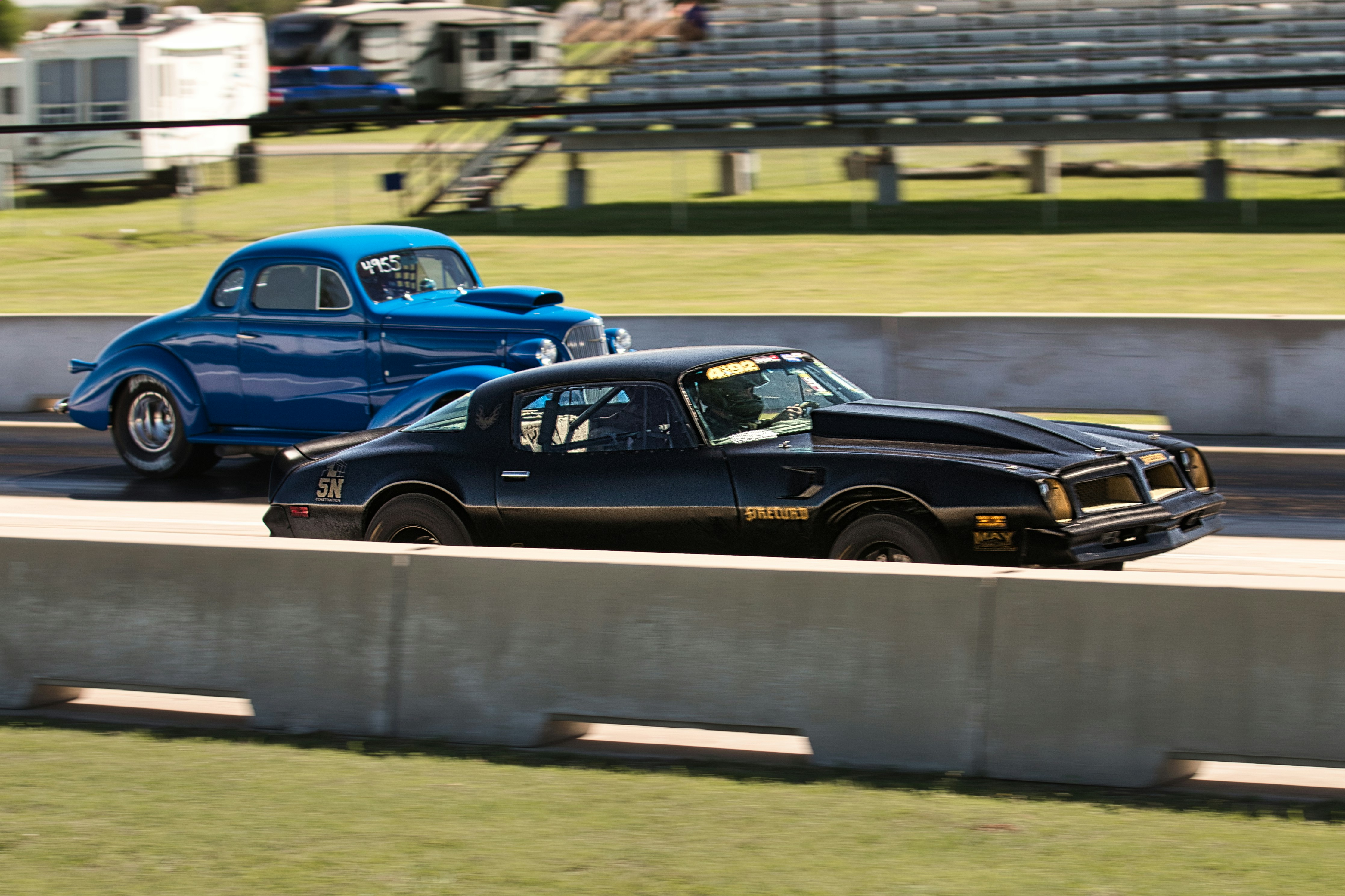 Classic cars competing on a drag strip with empty bleachers in the background.