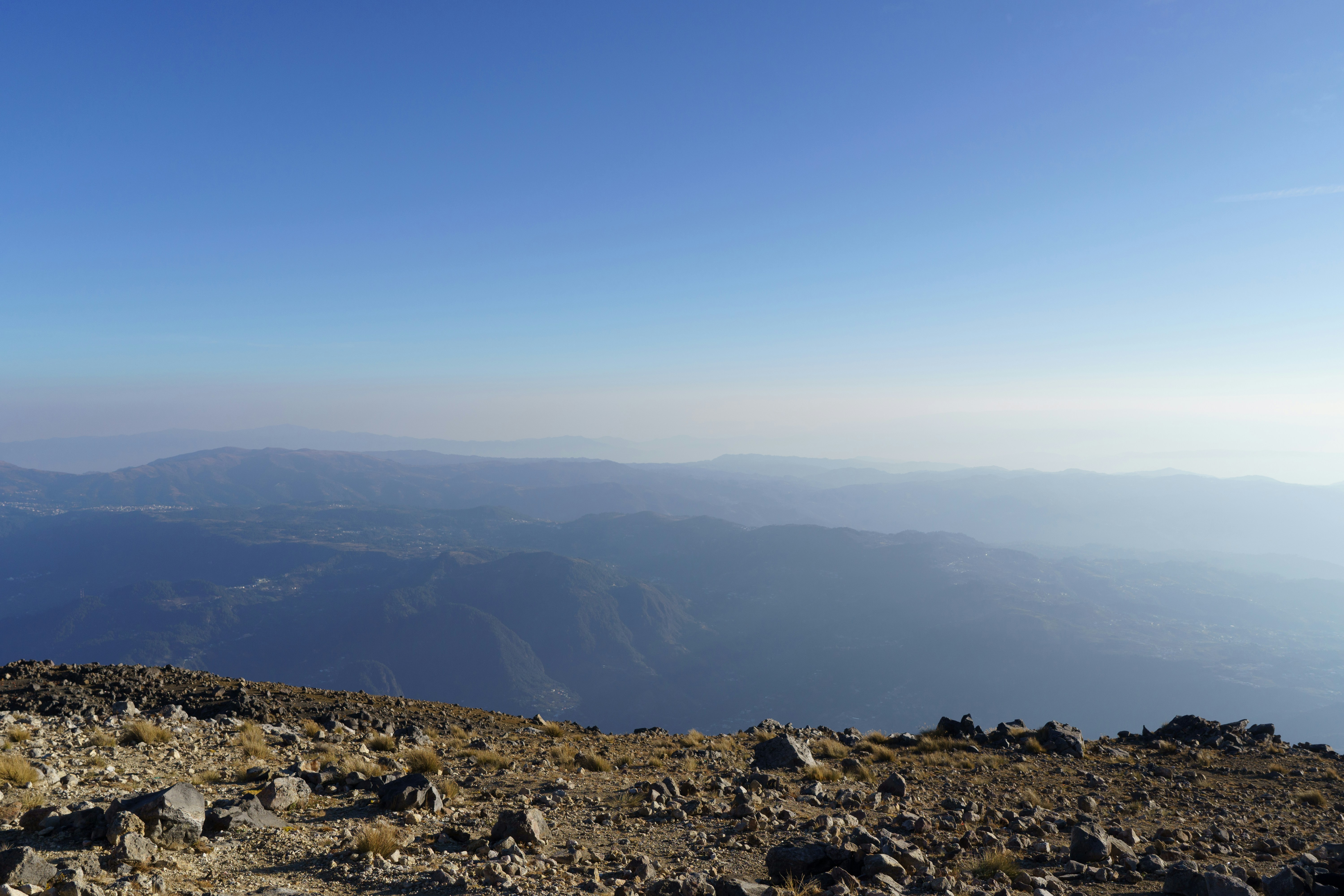 Person standing atop a rocky mountain with expansive views of distant, hazy peaks under a clear blue sky.