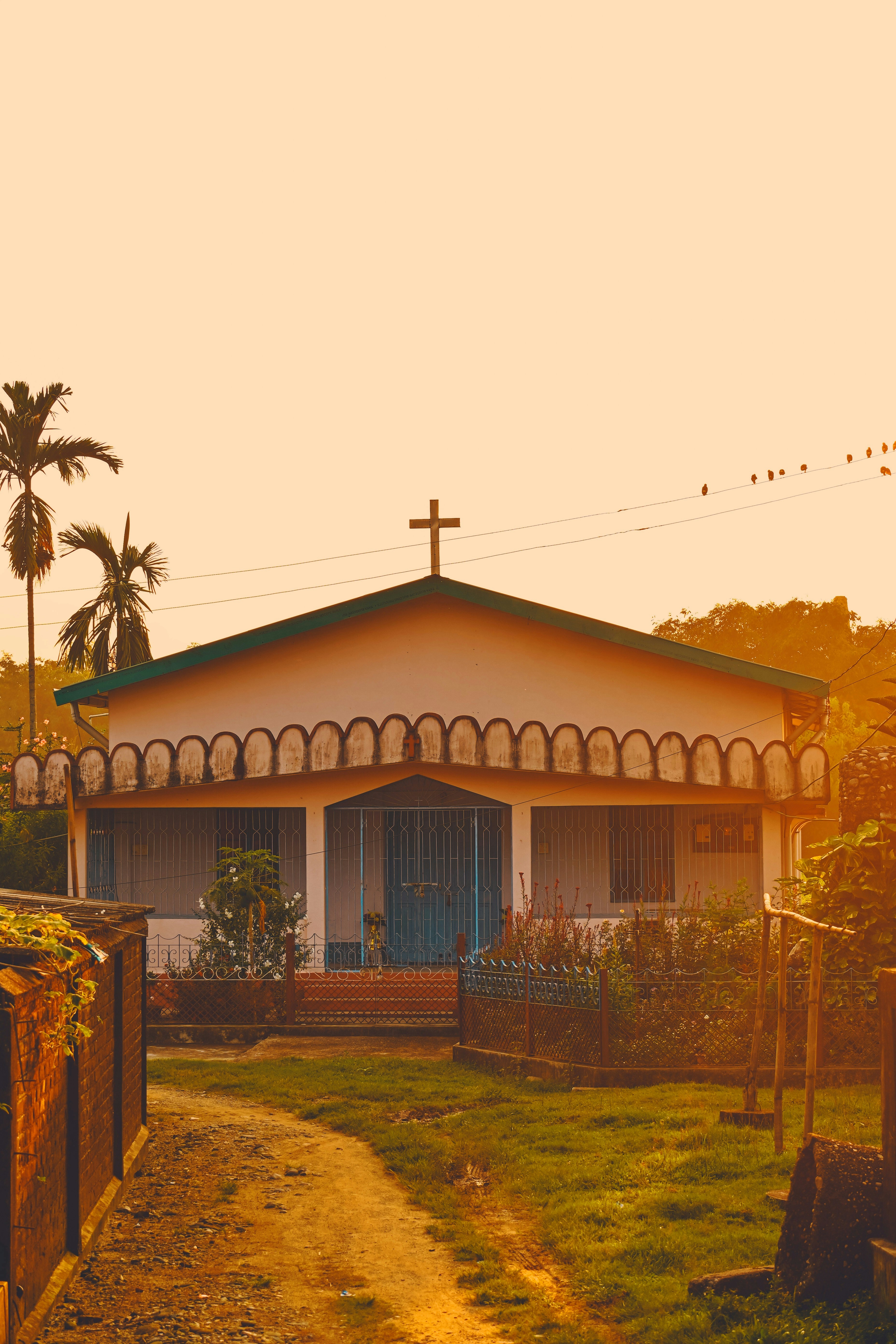 A serene church with a cross atop, surrounded by lush greenery and a winding path, illuminated by the warm glow of sunset.