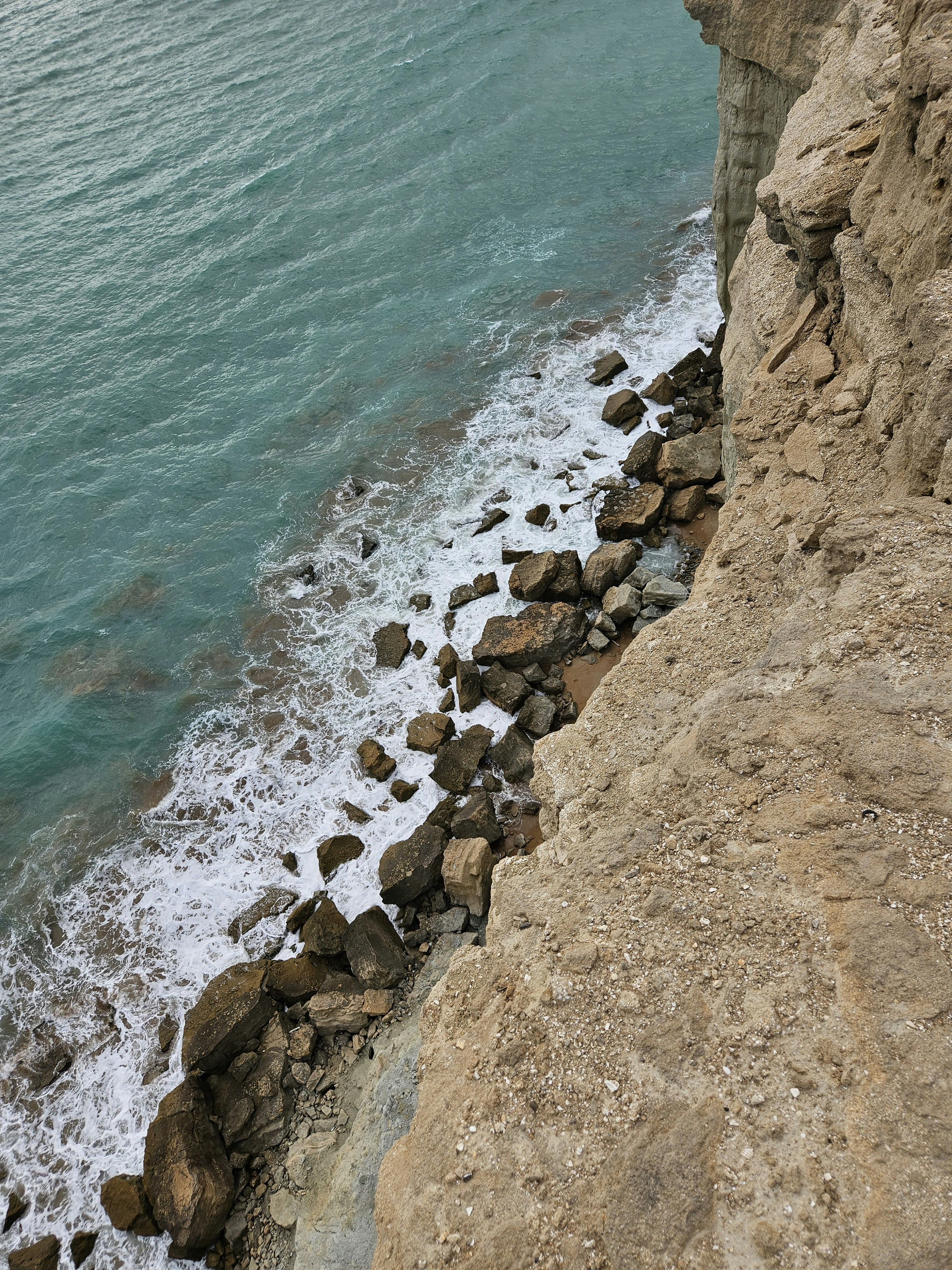 Rugged cliff edge flanks a rocky shoreline where turquoise waves crash against stones, highlighting weathered textures and erosion patterns.