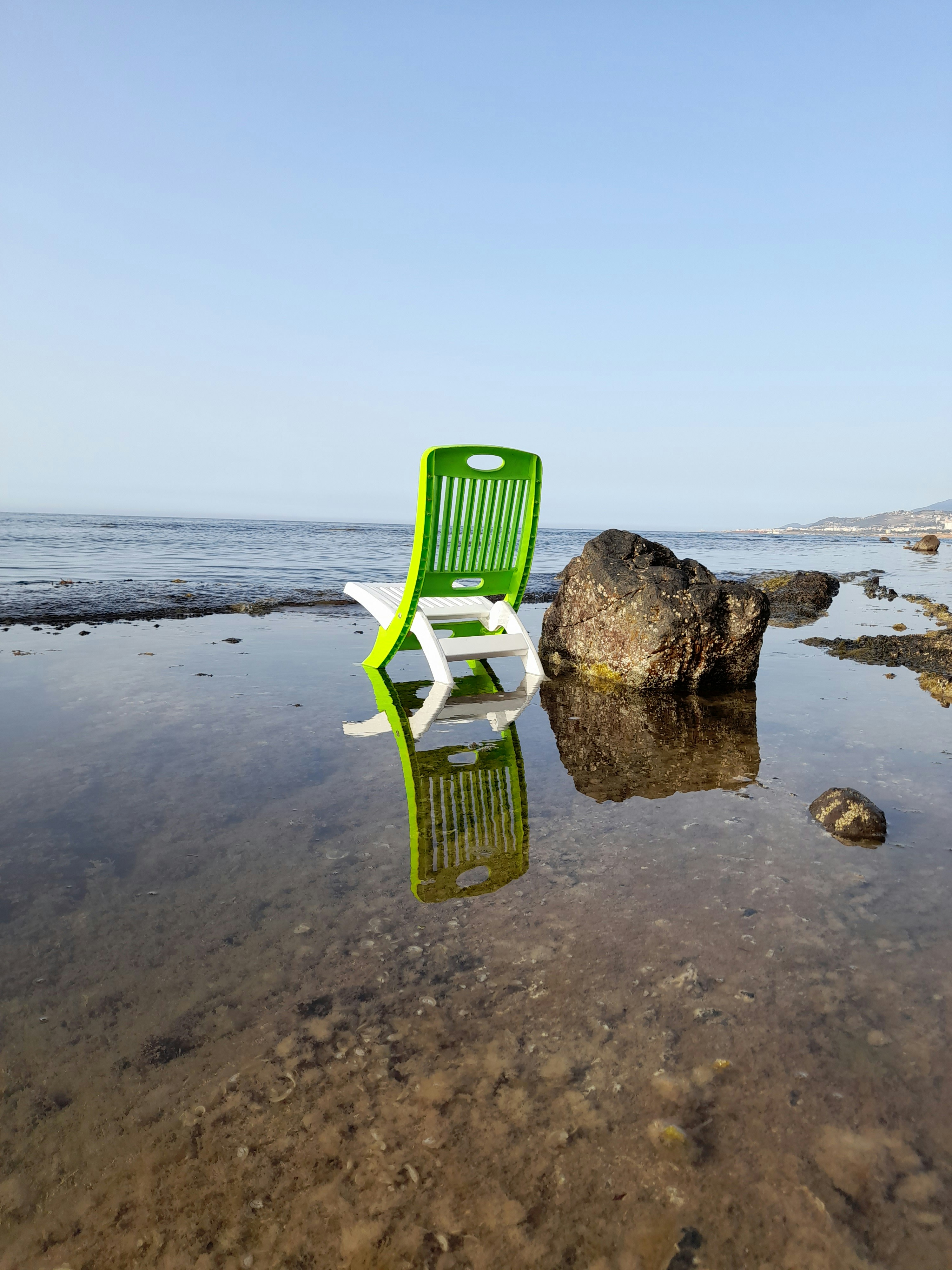Bright green plastic chair rests in a shallow rock pool, its reflection mirrored on the glassy water as a calm sea and distant rocks frame the scene.