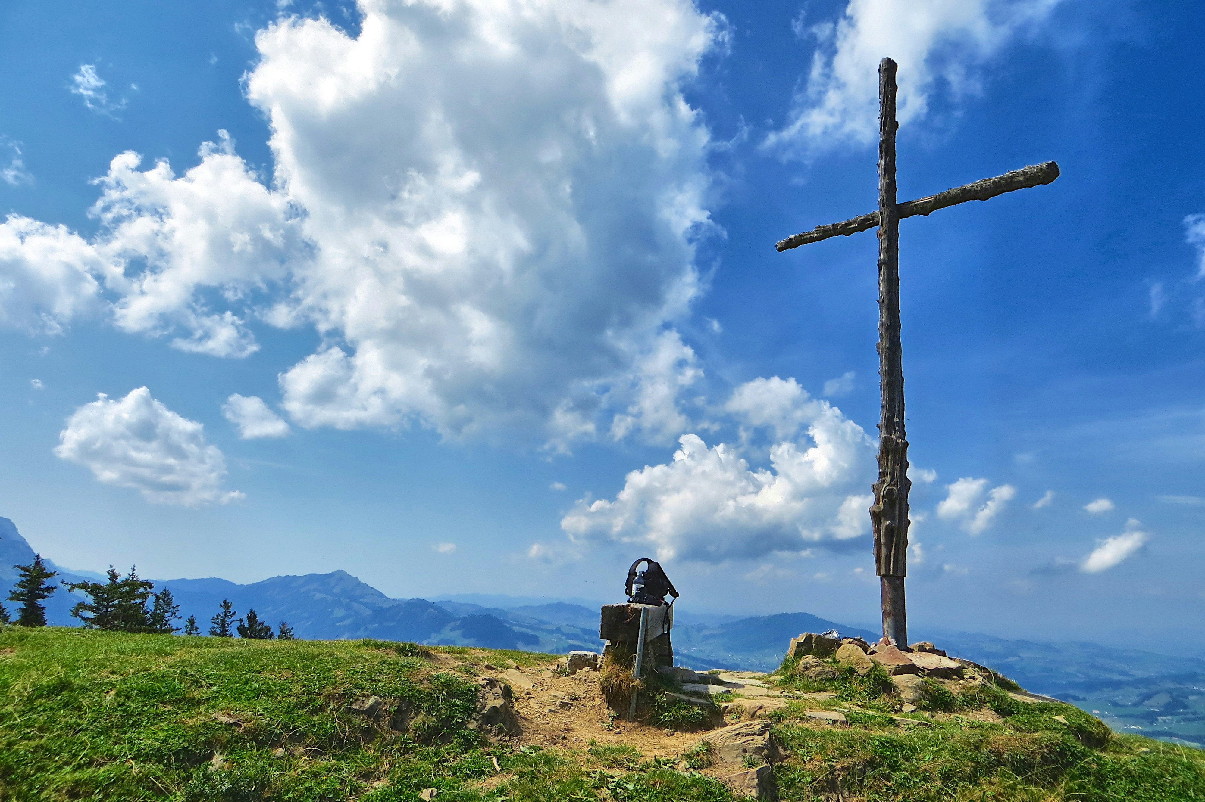 Photograph of a lone cross on a grassy summit with a kneeling figure nearby and distant mountains under a bright blue sky.
