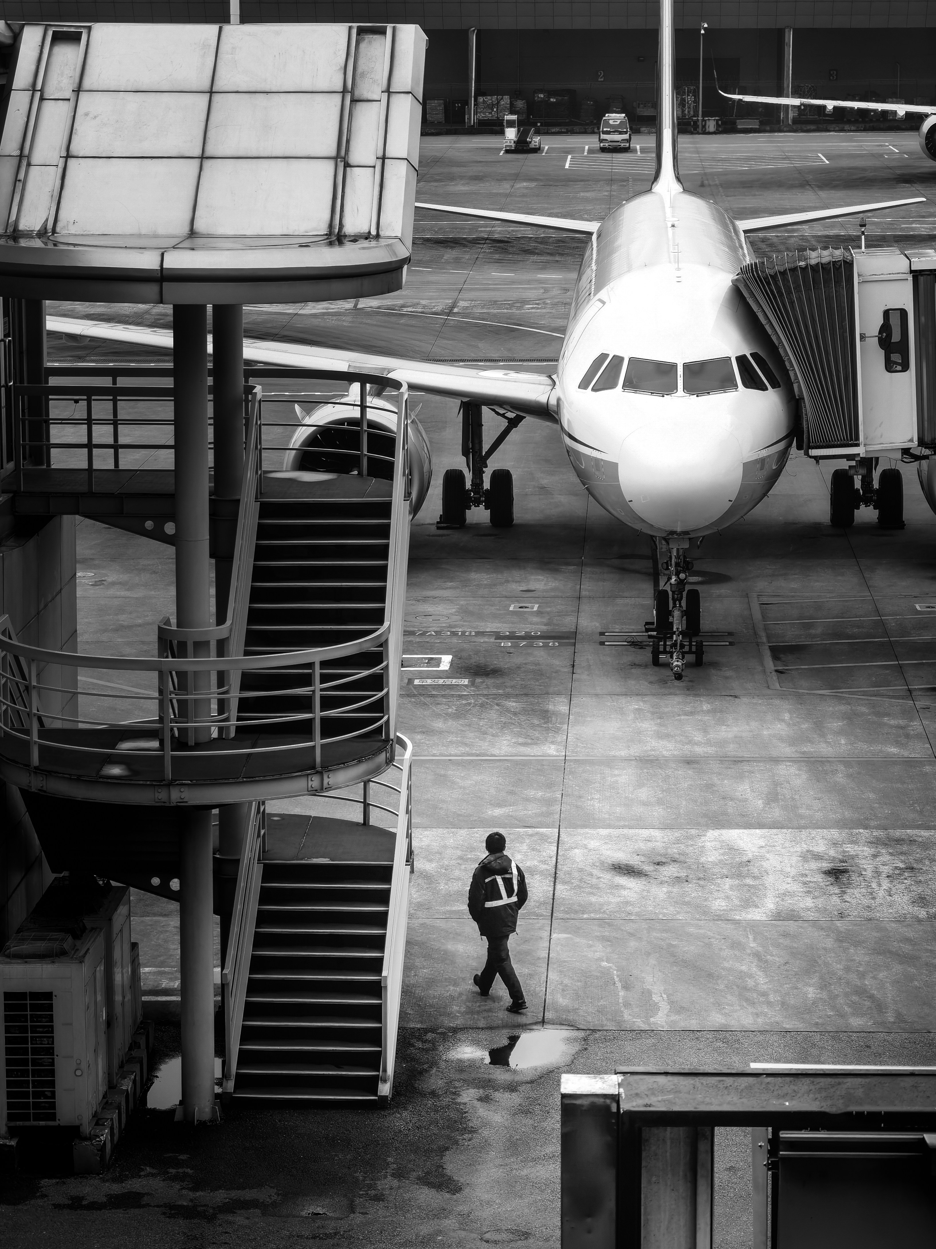 Monochrome photograph of an airplane at the gate with a ground crew member walking along the tarmac beside a jet bridge.