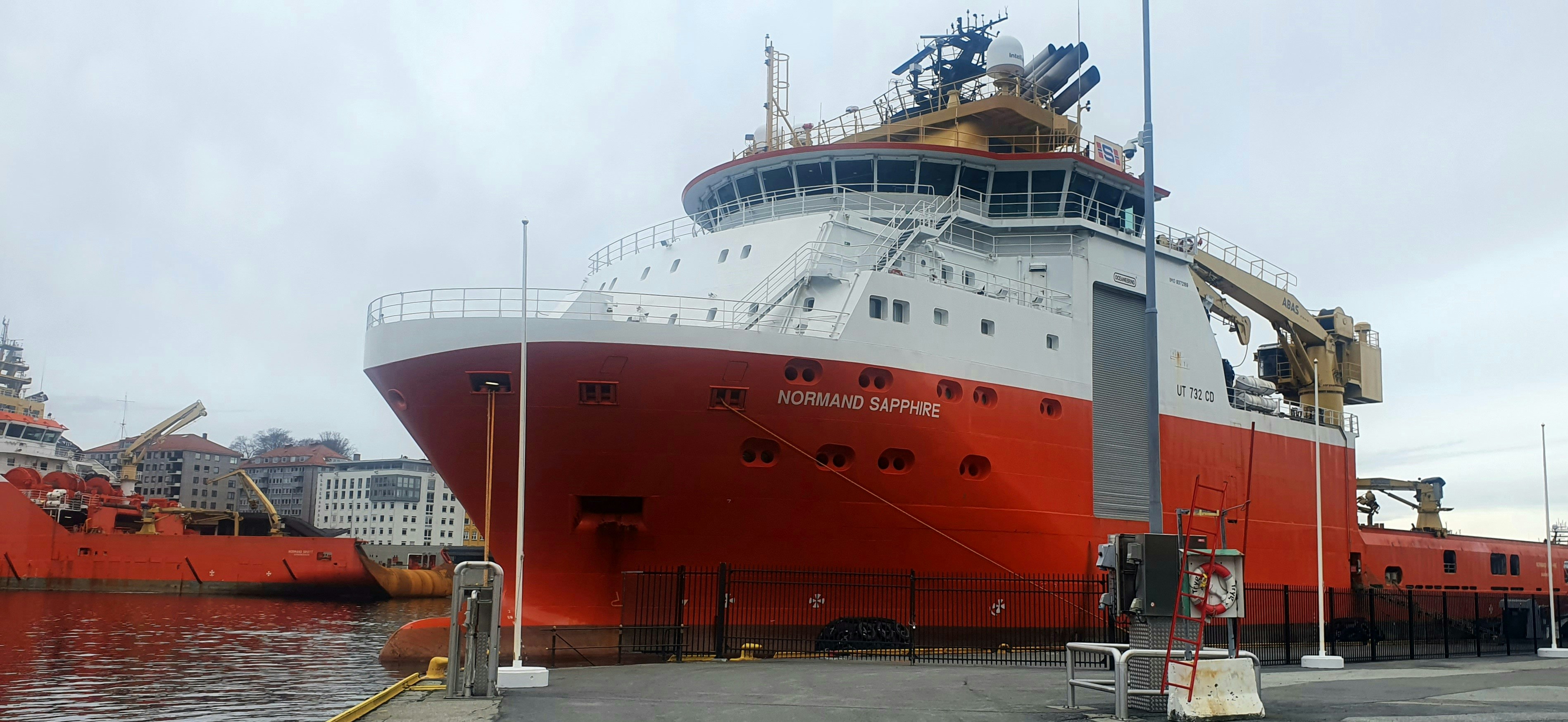 Large offshore support vessel, Normand Sapphire, docked beside other ships in a bustling harbor. Industrial structures and cloudy sky form the backdrop.